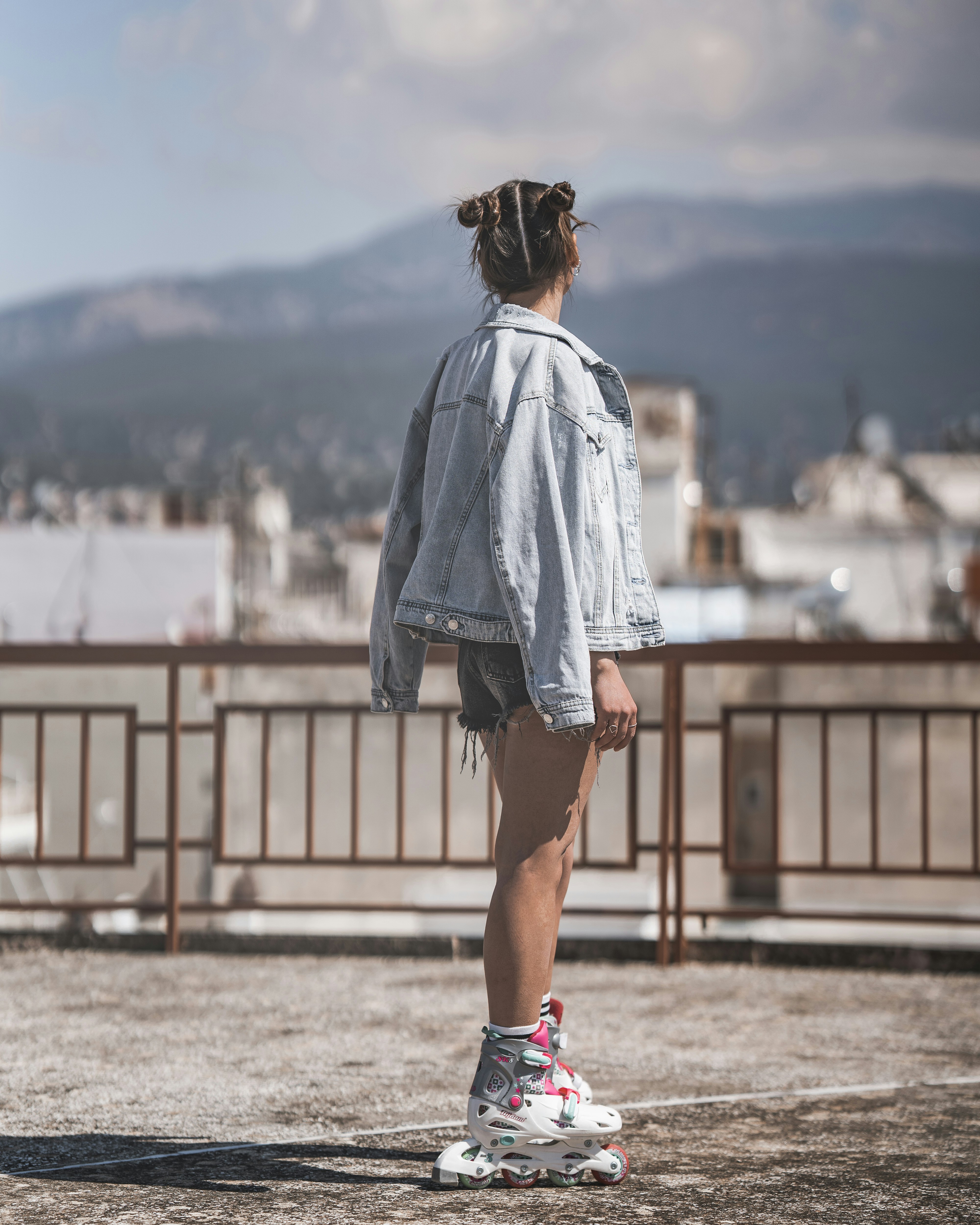 A rollerblader in a denim jacket and shorts stands on a rooftop, gazing at the distant mountains under a clear sky.