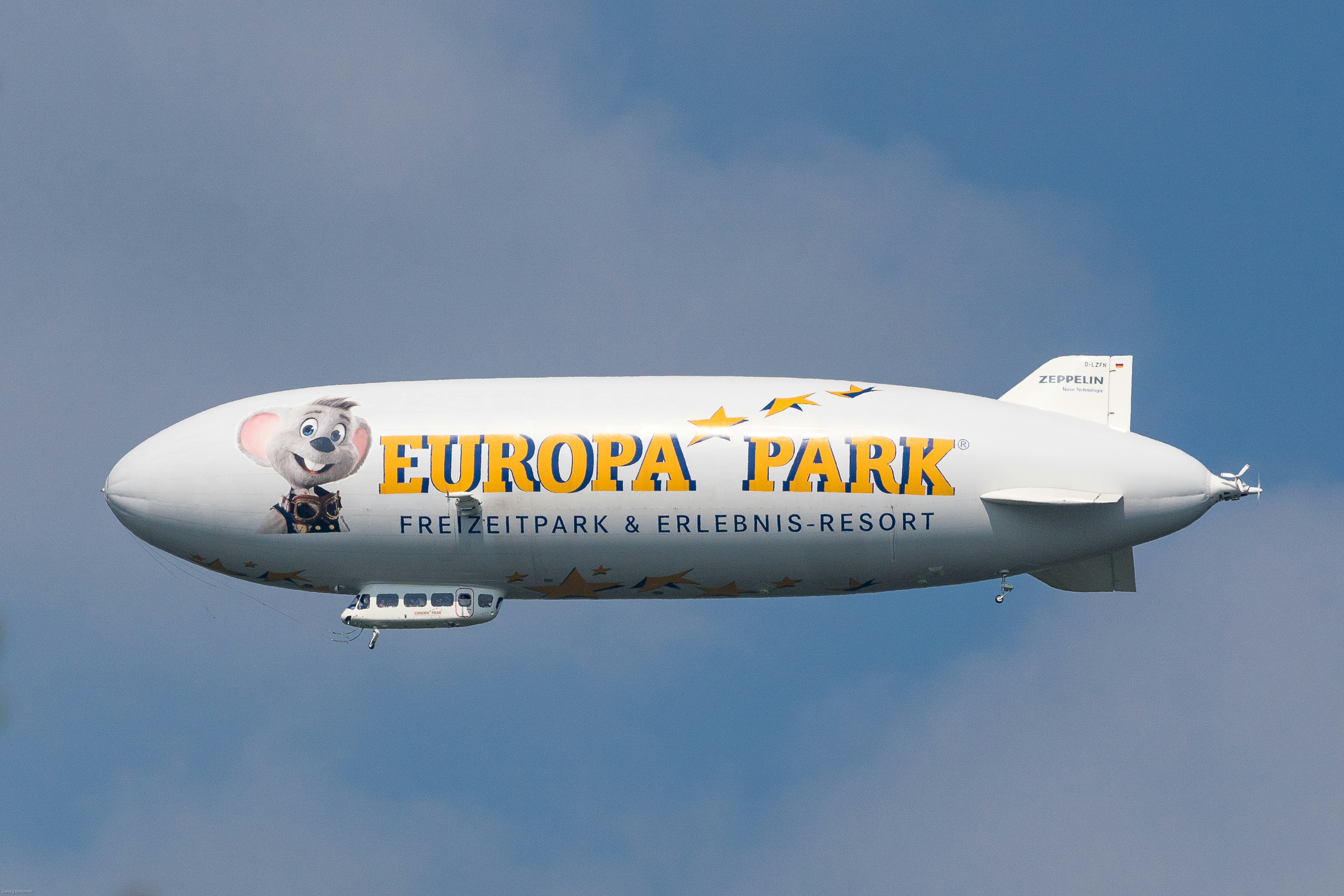 A blimp advertising Europa Park glides through a blue sky, showcasing its vibrant branding and playful mascot. The scene captures the essence of leisure and adventure.
