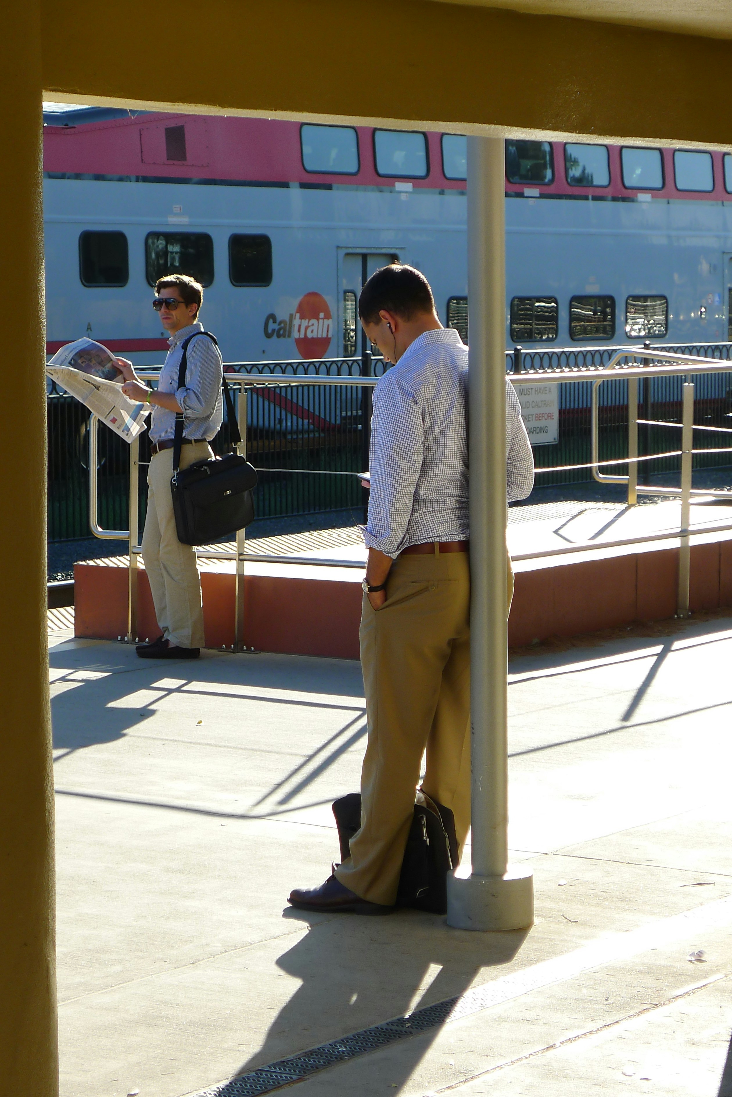 Two commuters await their train at a station, one reading a newspaper while the other stands pensively, framed by architectural elements.