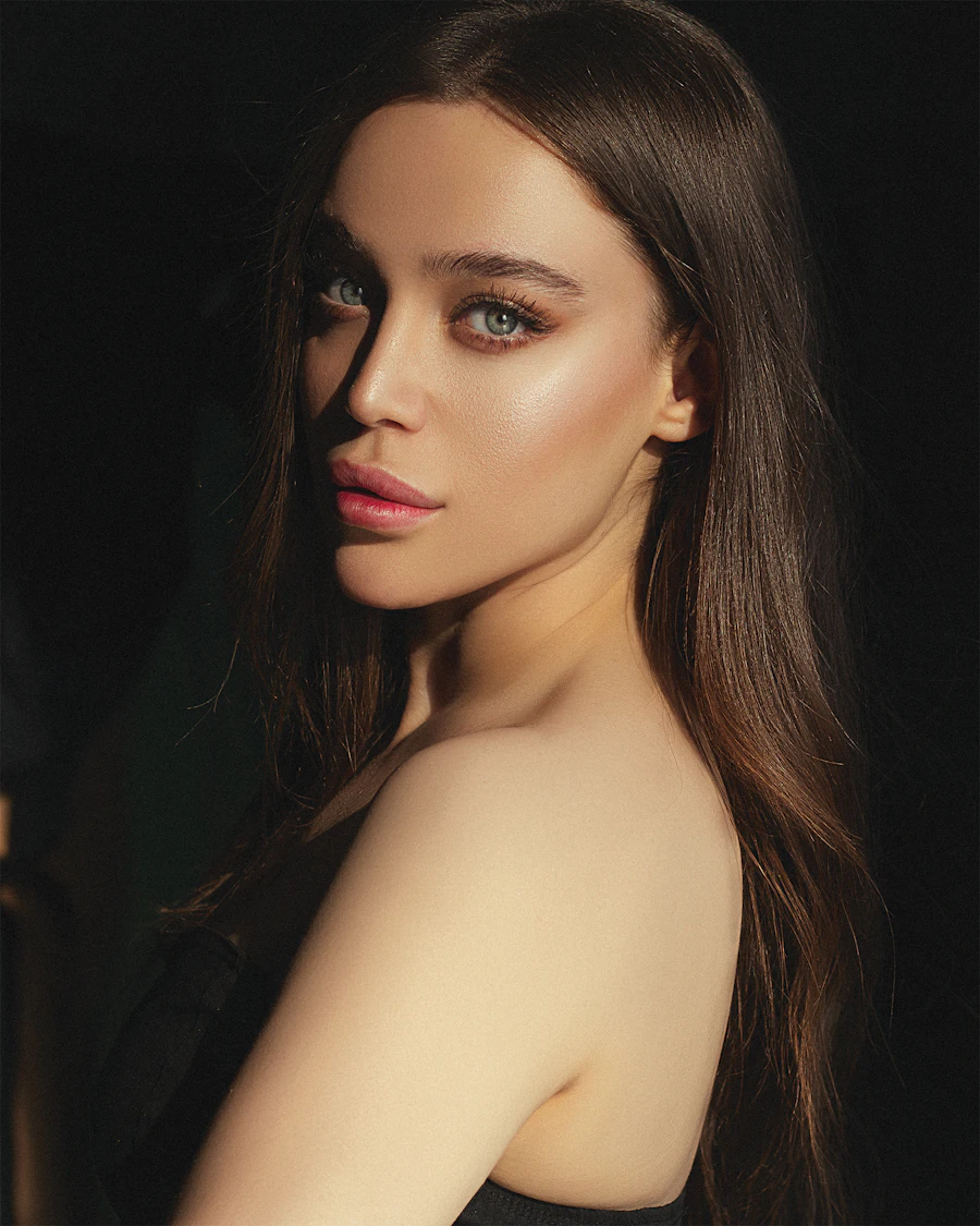 Young woman with long brown hair posed against a dark background, lit by warm directional light that highlights her face and shoulder. She looks toward the camera with a soft, serious expression.