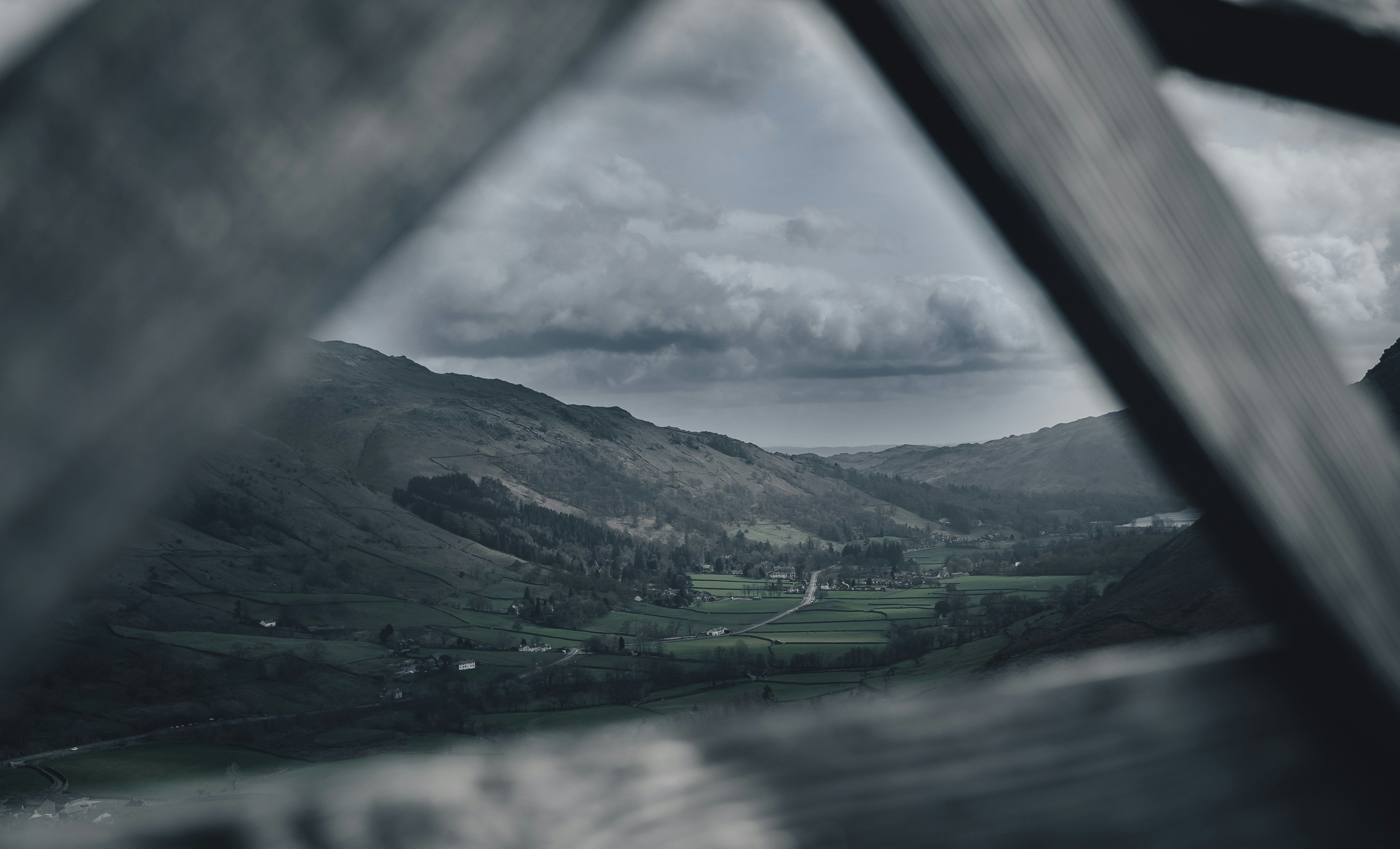 A panoramic view of a lush valley framed by wooden beams, showcasing rolling hills and a dramatic sky. The scene evokes a sense of tranquility and connection to nature.