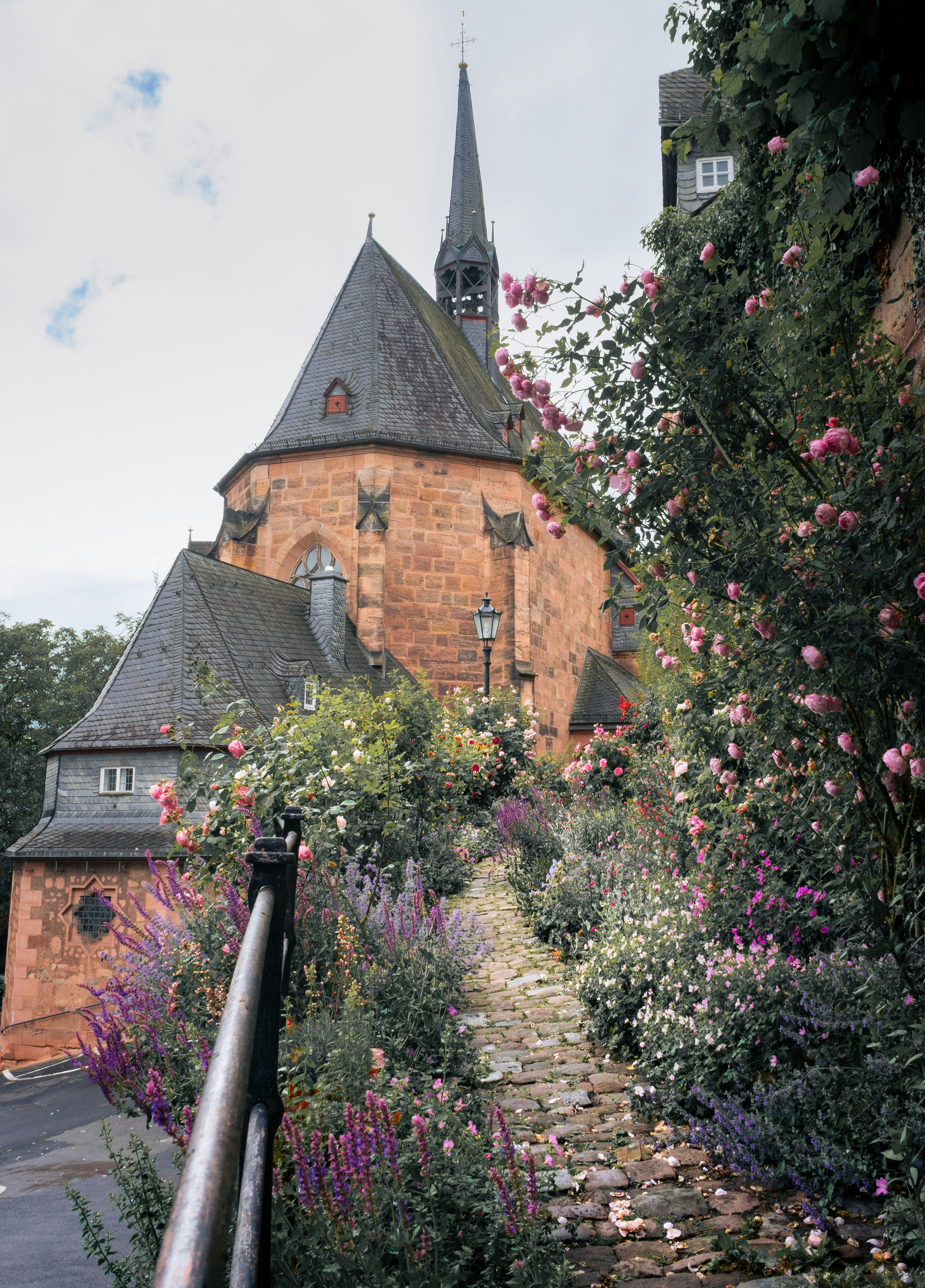 Charming pathway adorned with vibrant flowers leading to a historic stone church, framed by lush greenery. A quaint street lamp adds to the picturesque scene.
