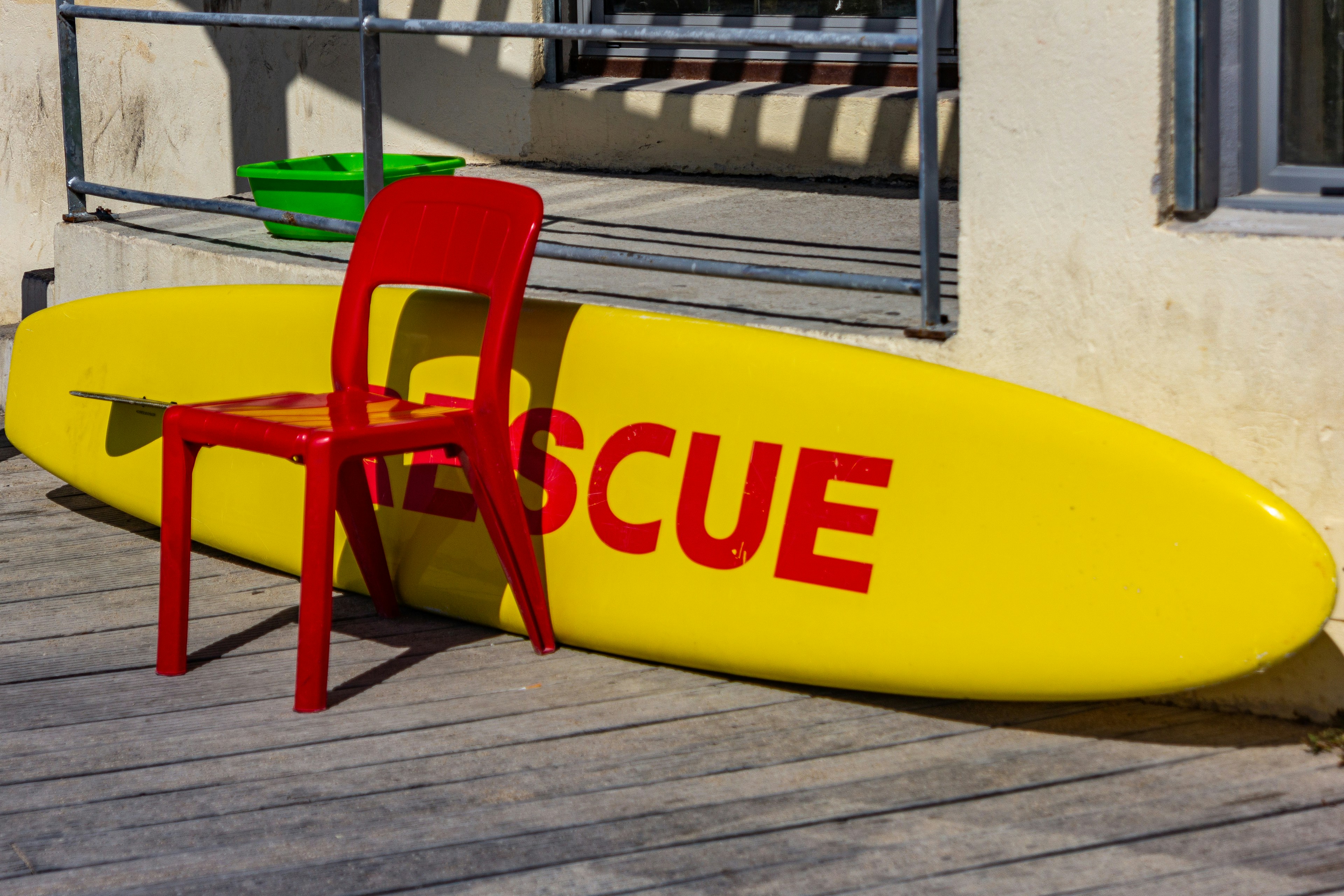 Yellow rescue surfboard with red lettering leaning against a wall, accompanied by a red chair on a wooden deck.