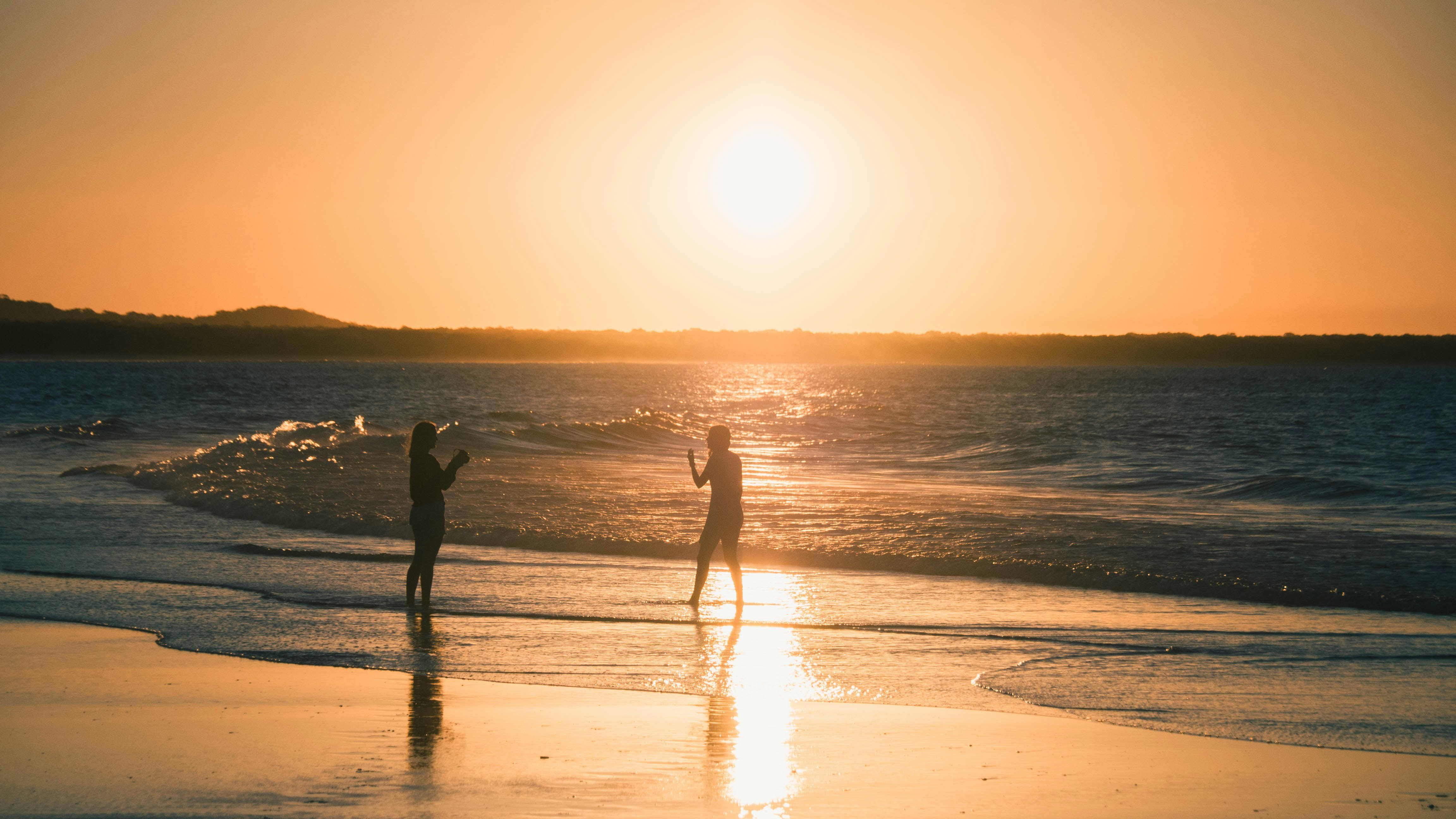 Two figures capturing memories on a sunlit beach as the sun sets on the horizon.