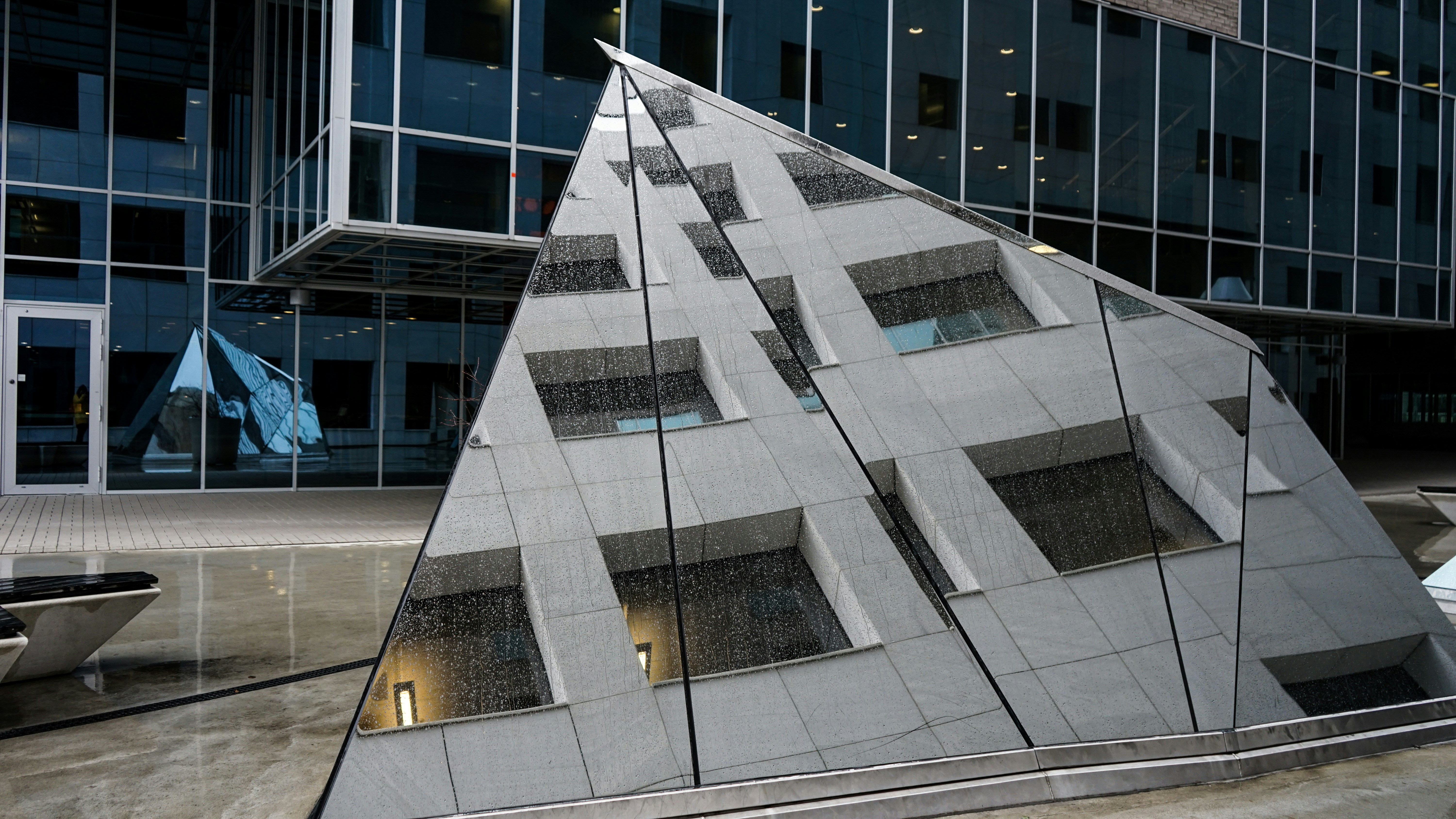 Abstract architectural sculpture reflecting the surrounding glass facade of a modern building. The interplay of light and geometry creates a dynamic visual narrative.