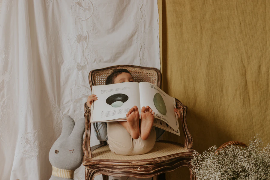 Child sitting in a wooden chair, absorbed in a large open book with playful illustrations.