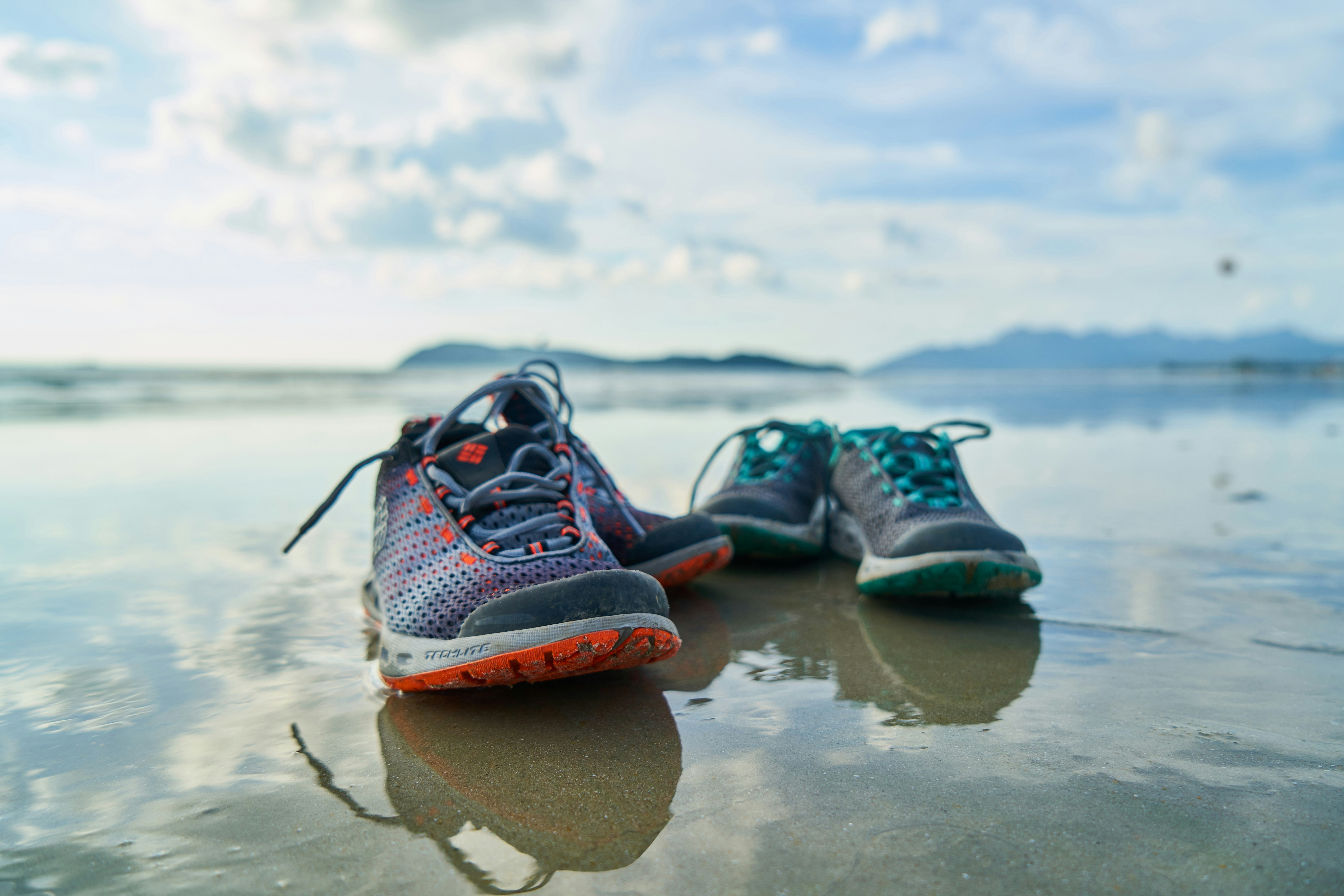 Three pairs of colorful shoes resting on a sandy beach, reflecting the sky and water. The scene evokes a sense of adventure and exploration.