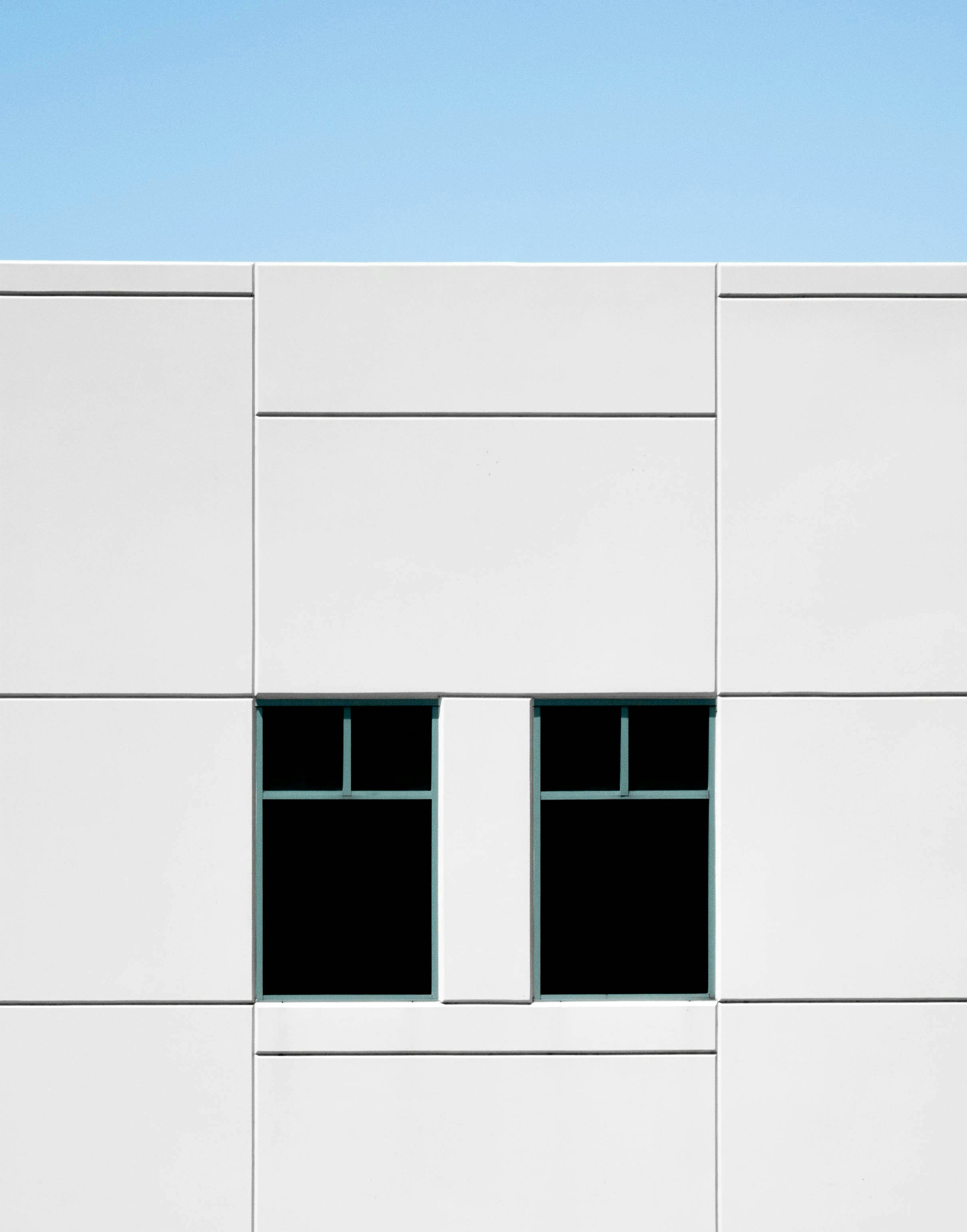 Two symmetrical windows set against a stark white wall, contrasting sharply with the clear blue sky above.