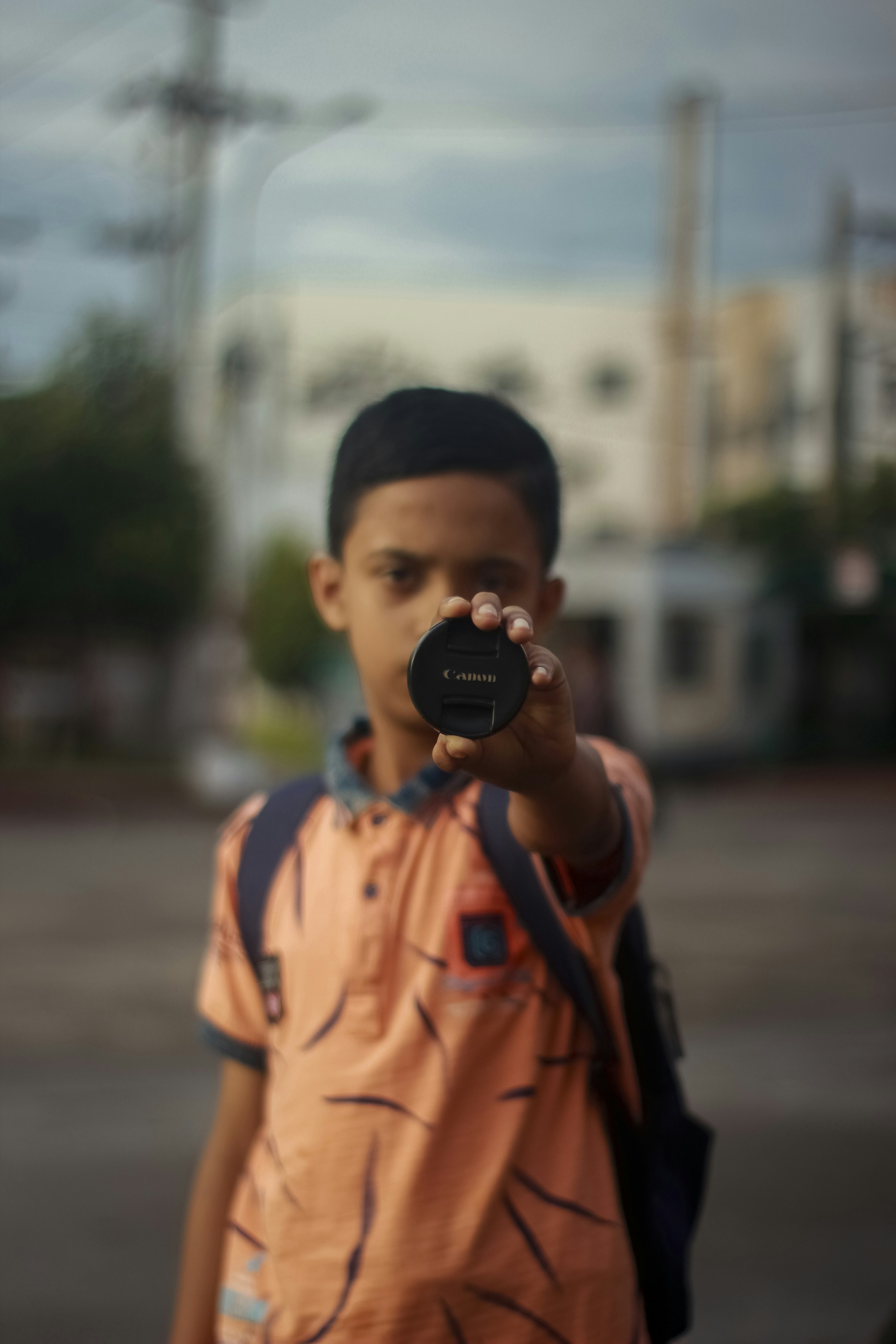 Boy holding a camera lens cap in front of him, with a blurred background of urban elements. 