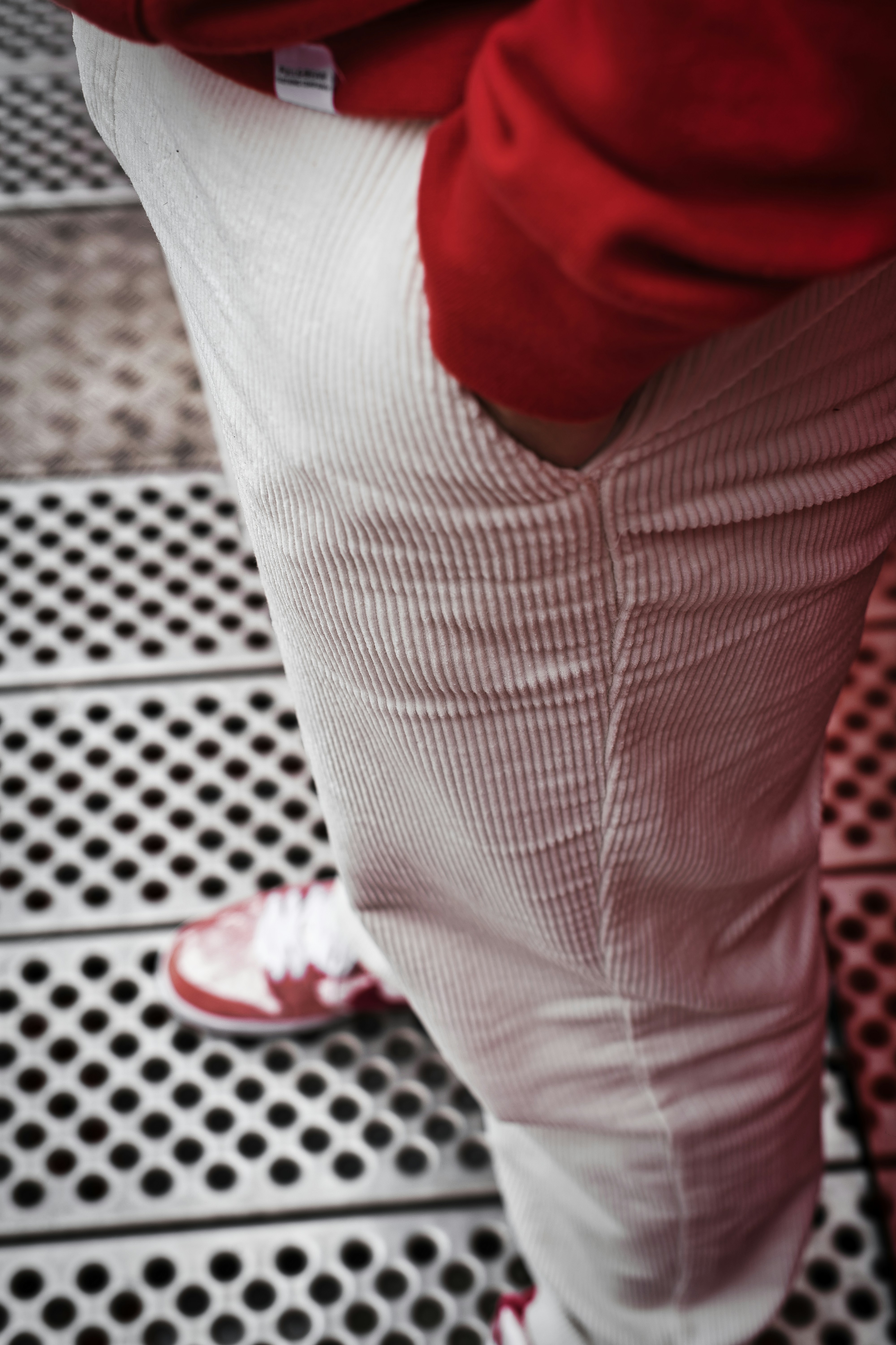 Close-up of a person wearing white corduroy pants and red sneakers, standing on a patterned metal surface.