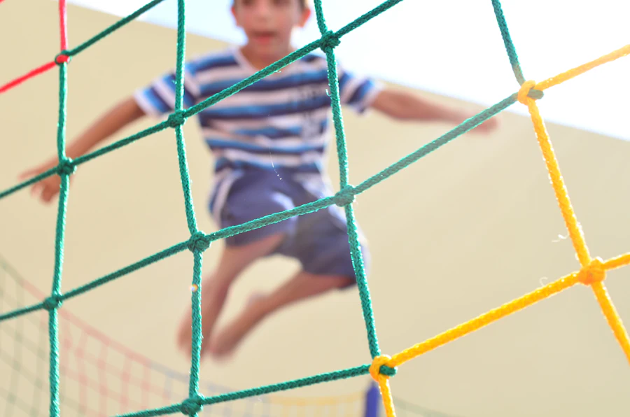 A child climbs a colorful playground net, legs dangling toward the camera, against a soft outdoor backdrop.