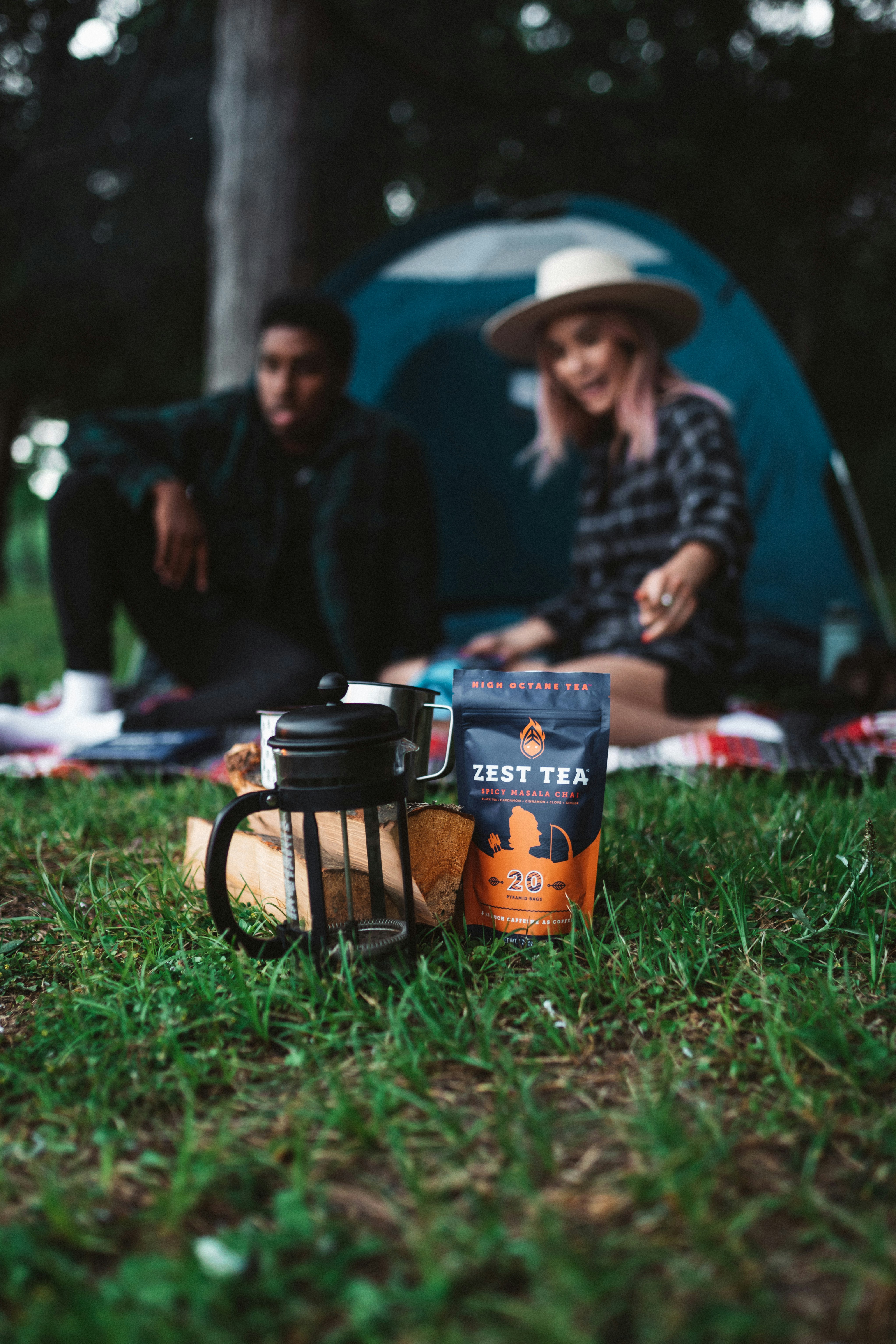 French press and a packet of Zest Tea on a picnic blanket in a campsite setting. Two individuals are enjoying the outdoors in the background.