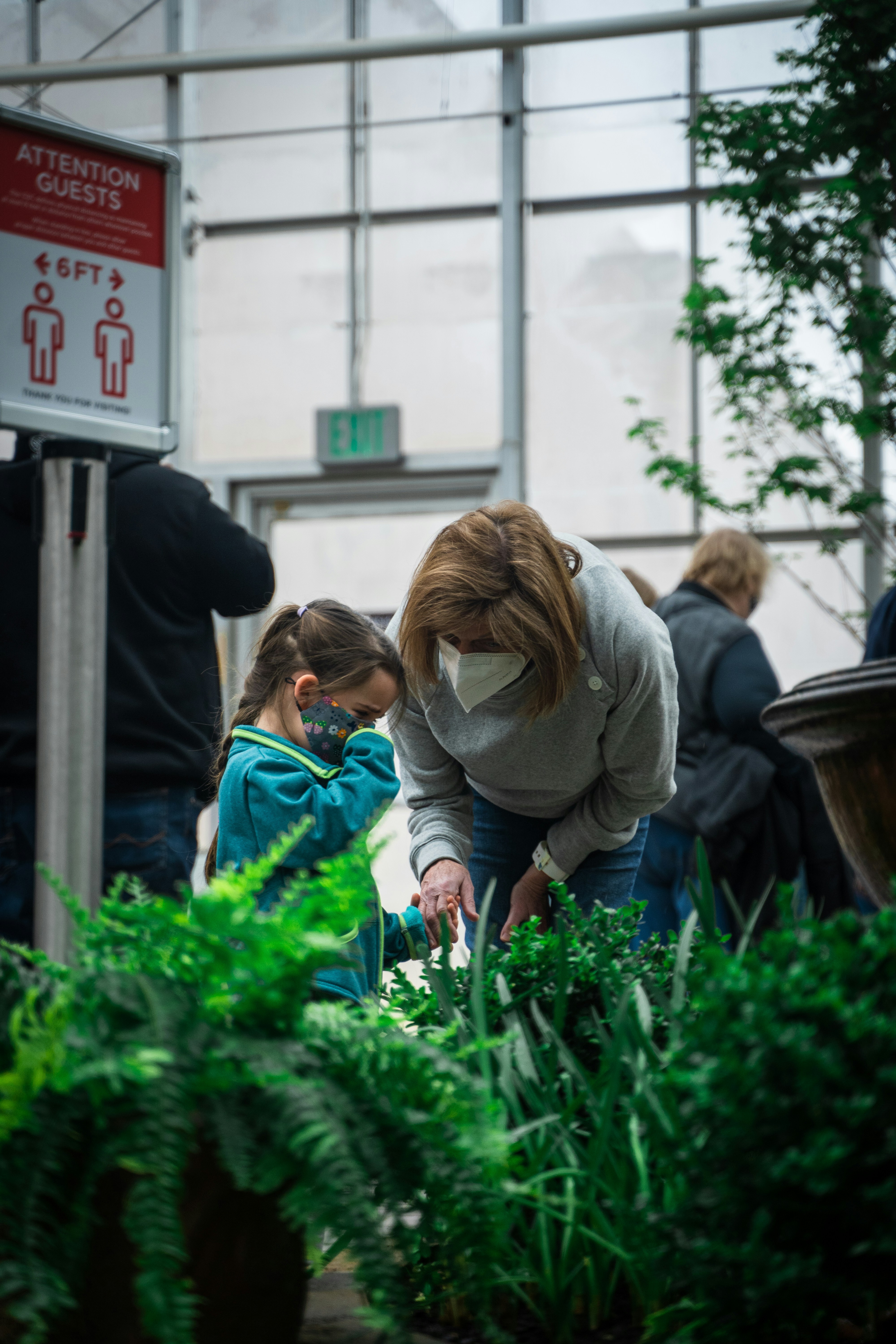A woman and a child attentively examine plants in a greenhouse, surrounded by lush greenery and fellow visitors. The scene captures a moment of shared curiosity and connection.