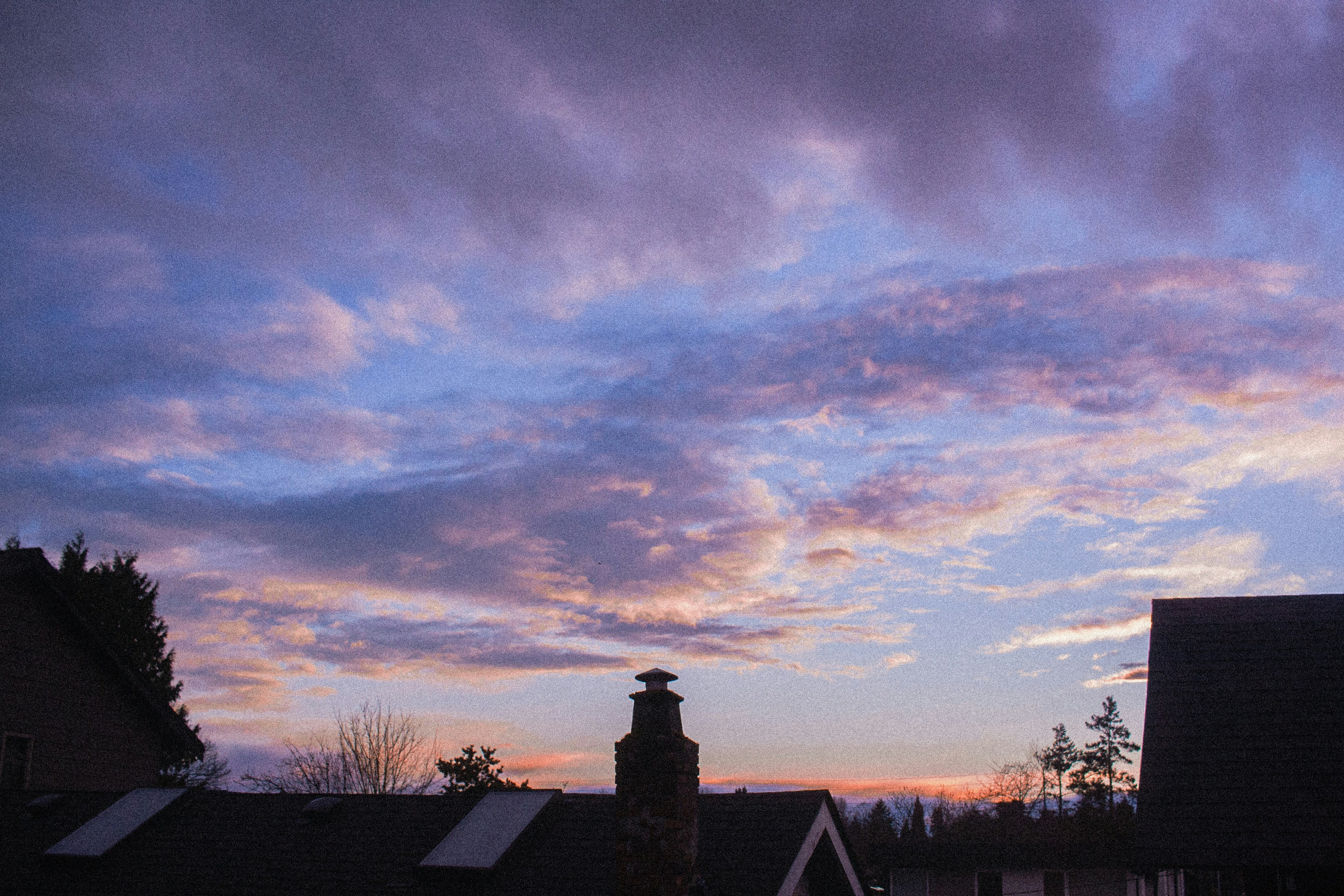 Vibrant twilight sky painted with hues of purple and orange, framed by silhouetted rooftops and chimney structures.