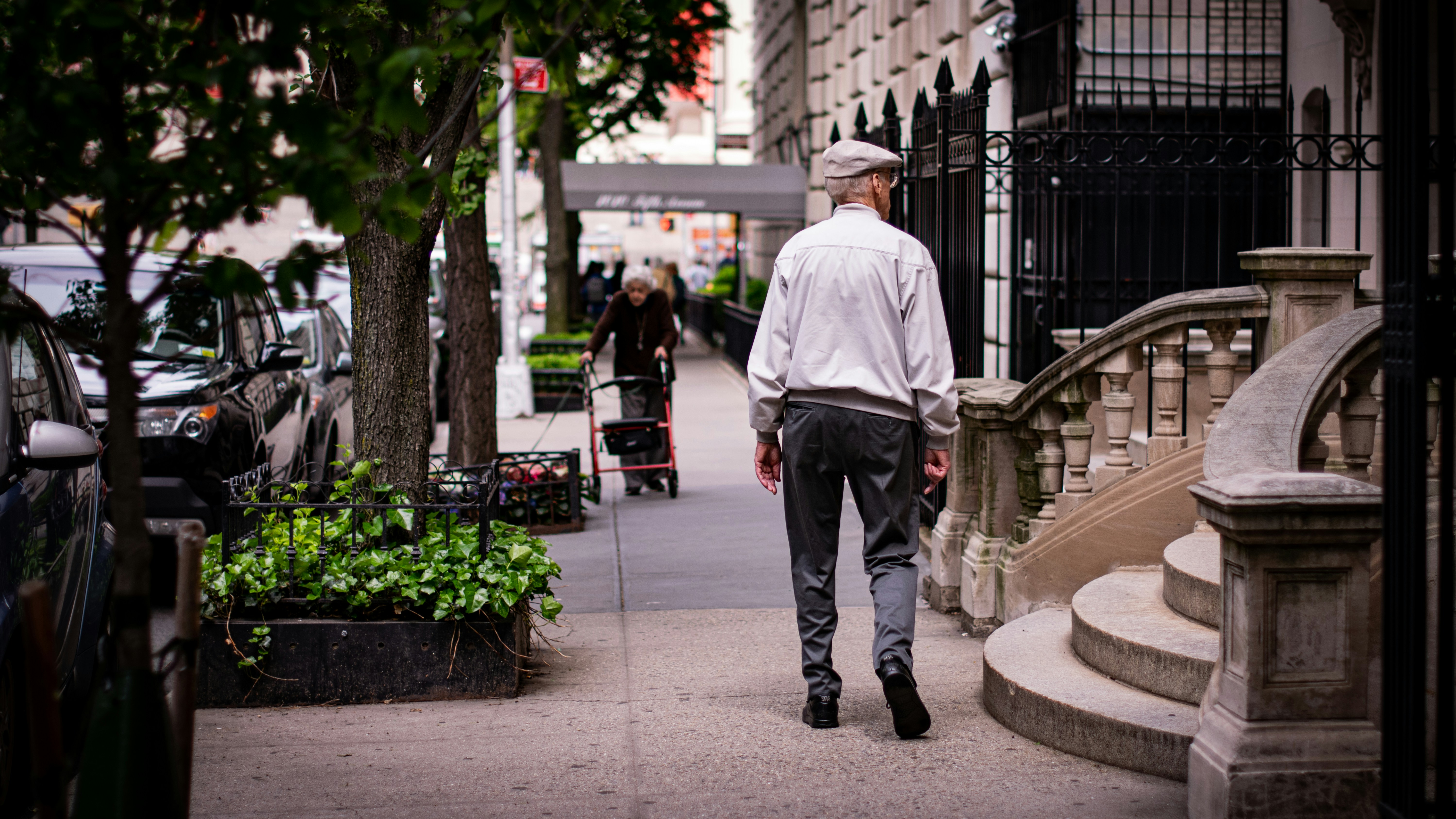 Elderly man walking down a city sidewalk, passing by lush greenery and elegant architecture. A woman with a cart is seen in the background.