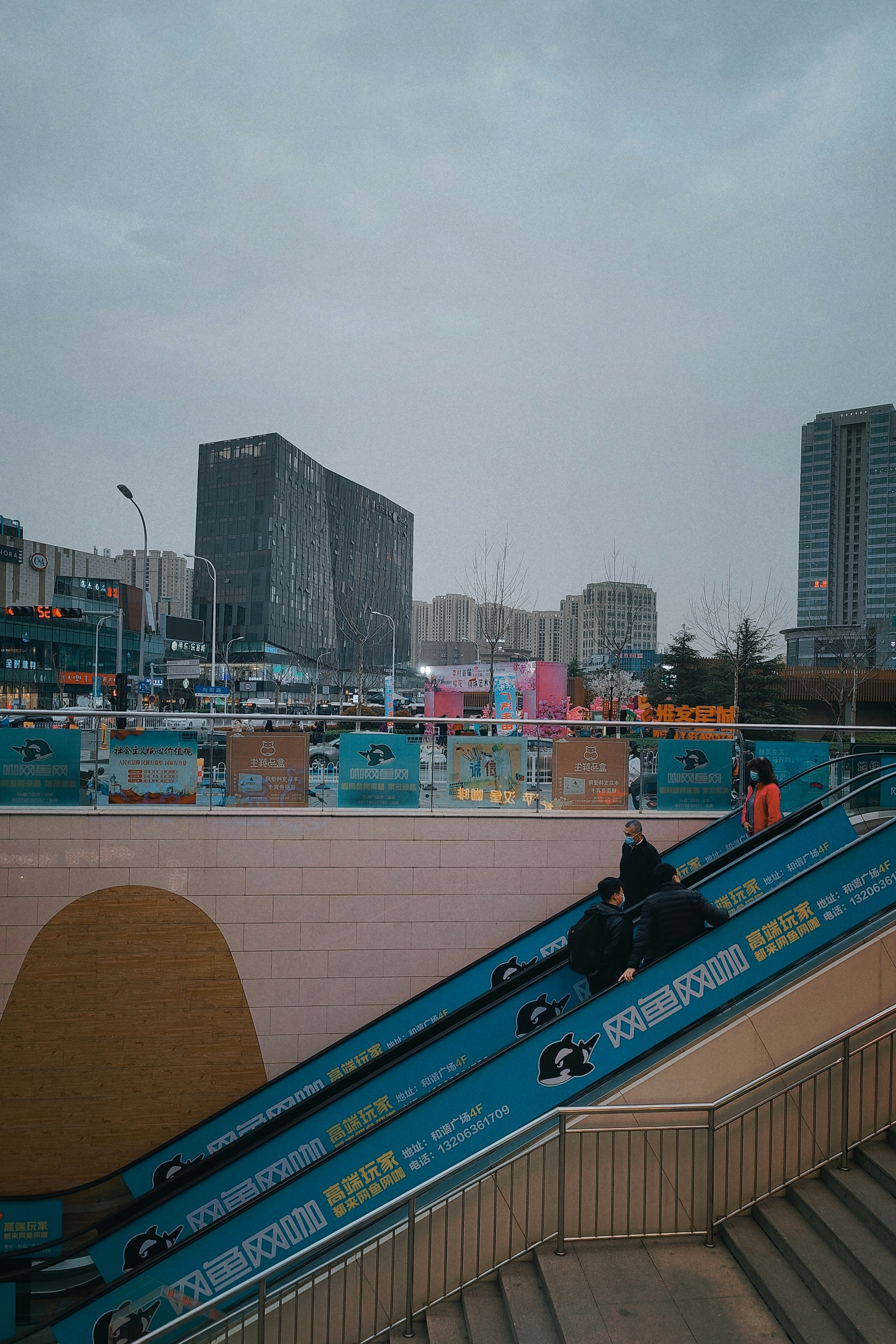 City plaza escalator lined with promotional blue banners as pedestrians move between levels under a cloudy skyline of modern high-rises.