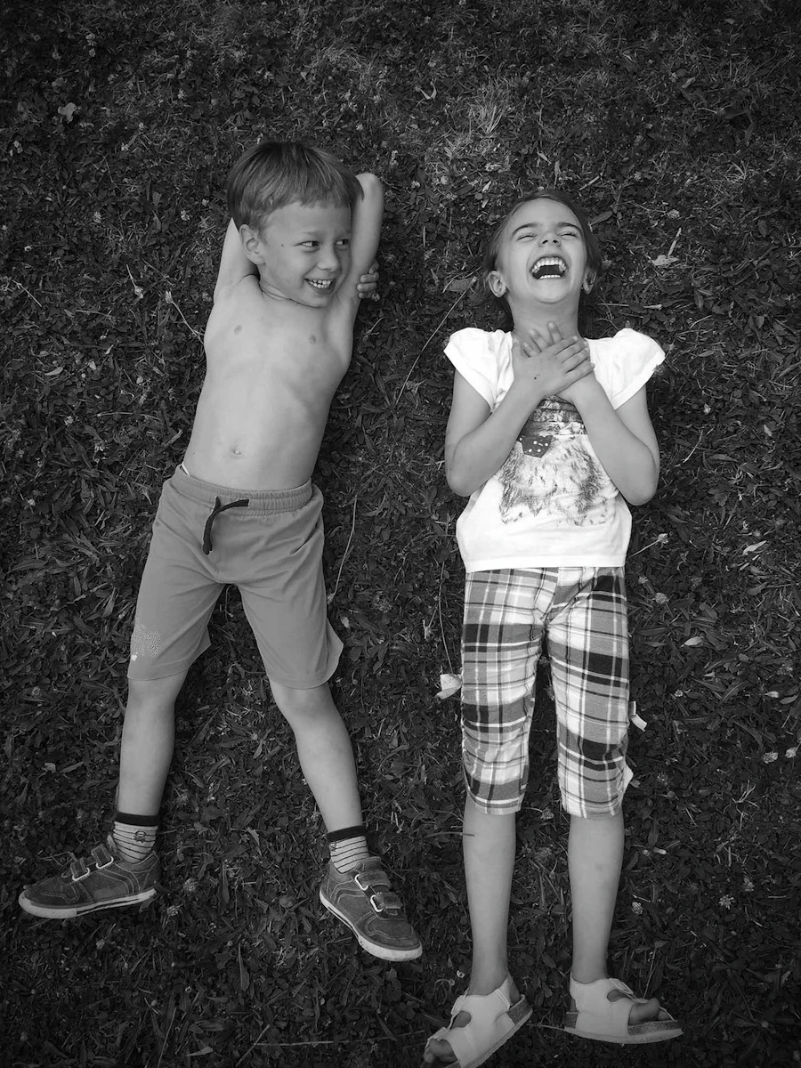 Two children lie on the grass, one shirtless boy and one girl in plaid shorts, both laughing with relaxed, carefree energy.