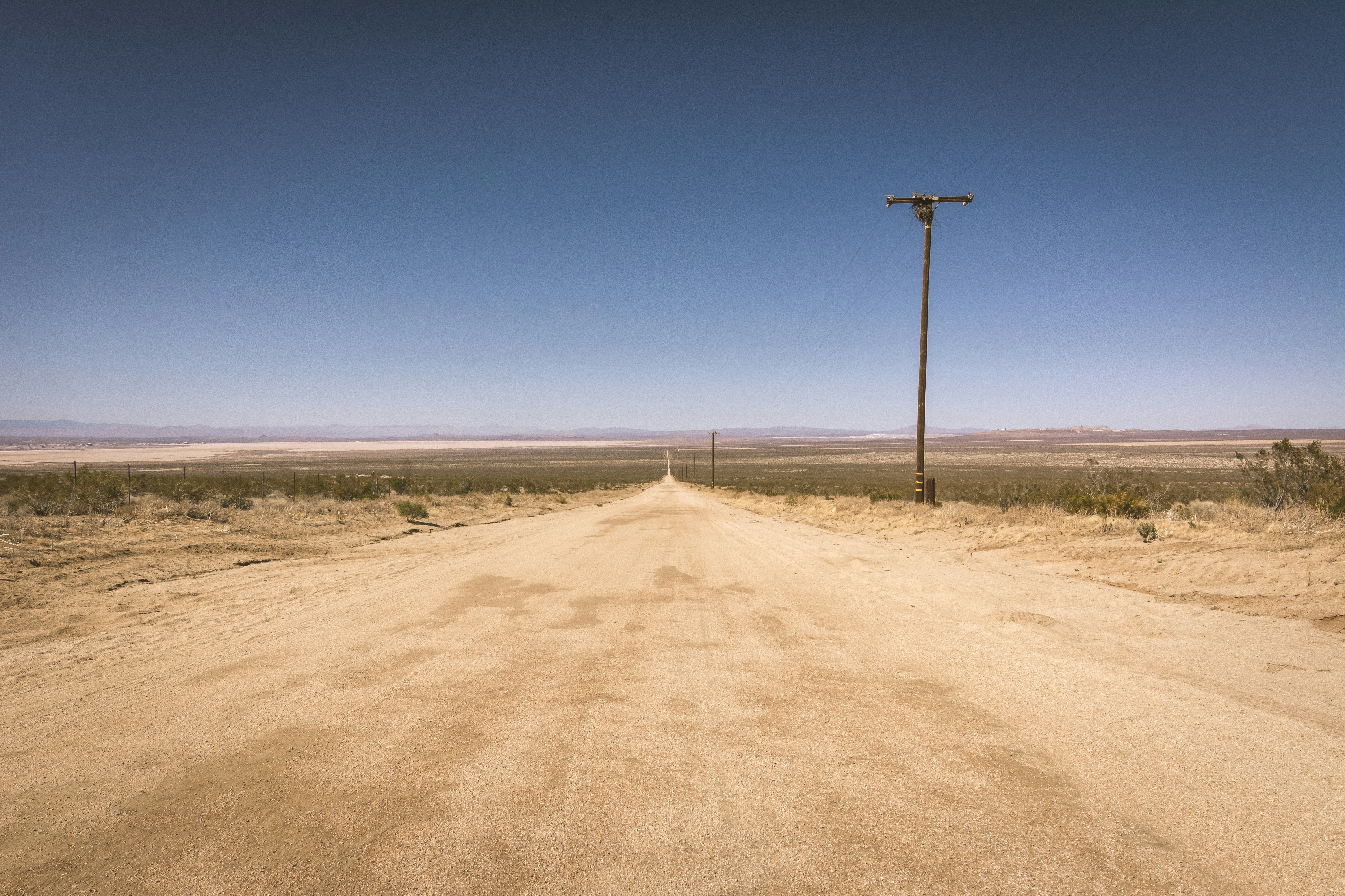 Lonely dirt road stretching into a vast desert landscape under a clear blue sky.