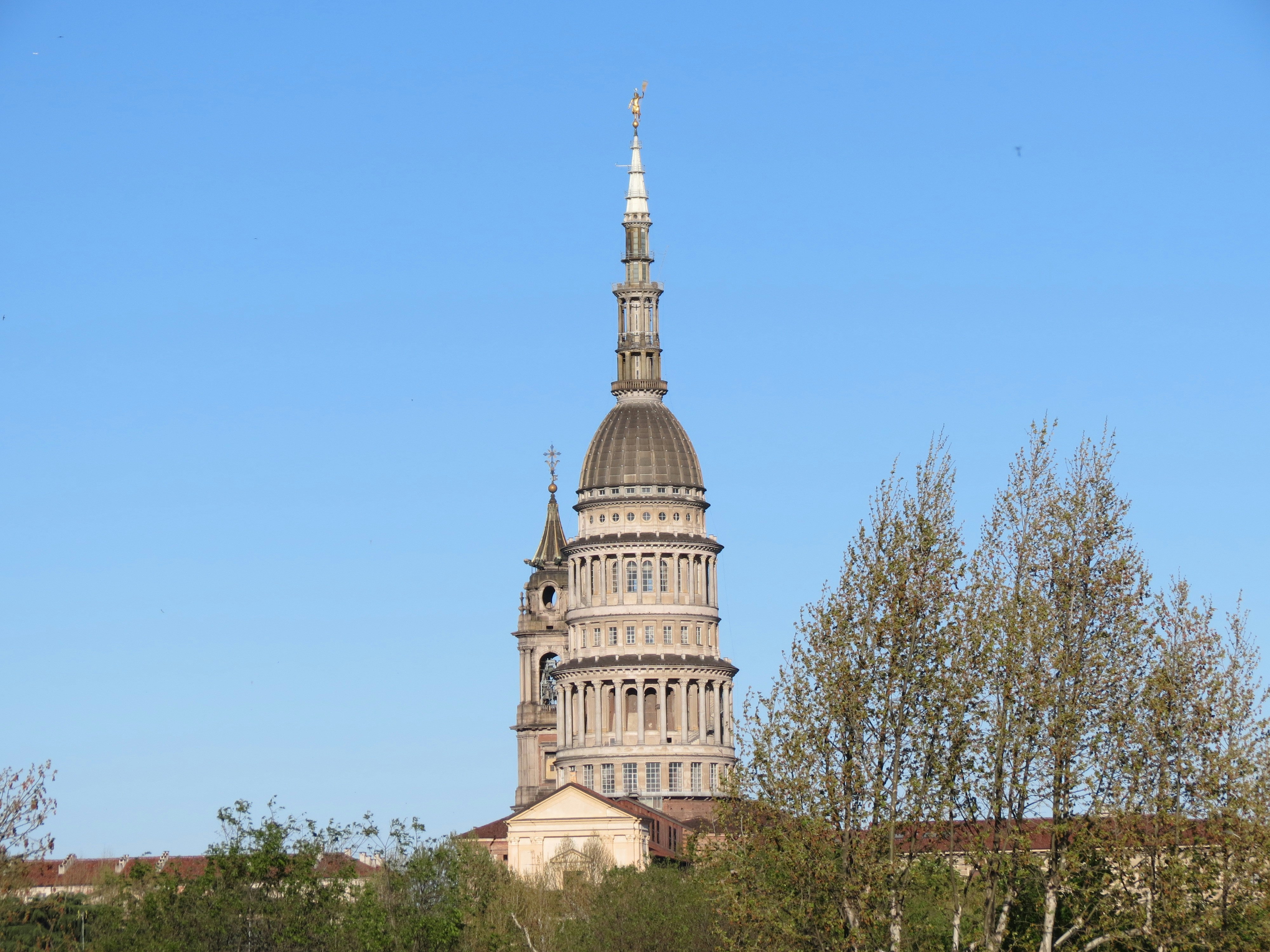 Tall architectural spire of a historic building rises against a clear blue sky, surrounded by lush greenery.