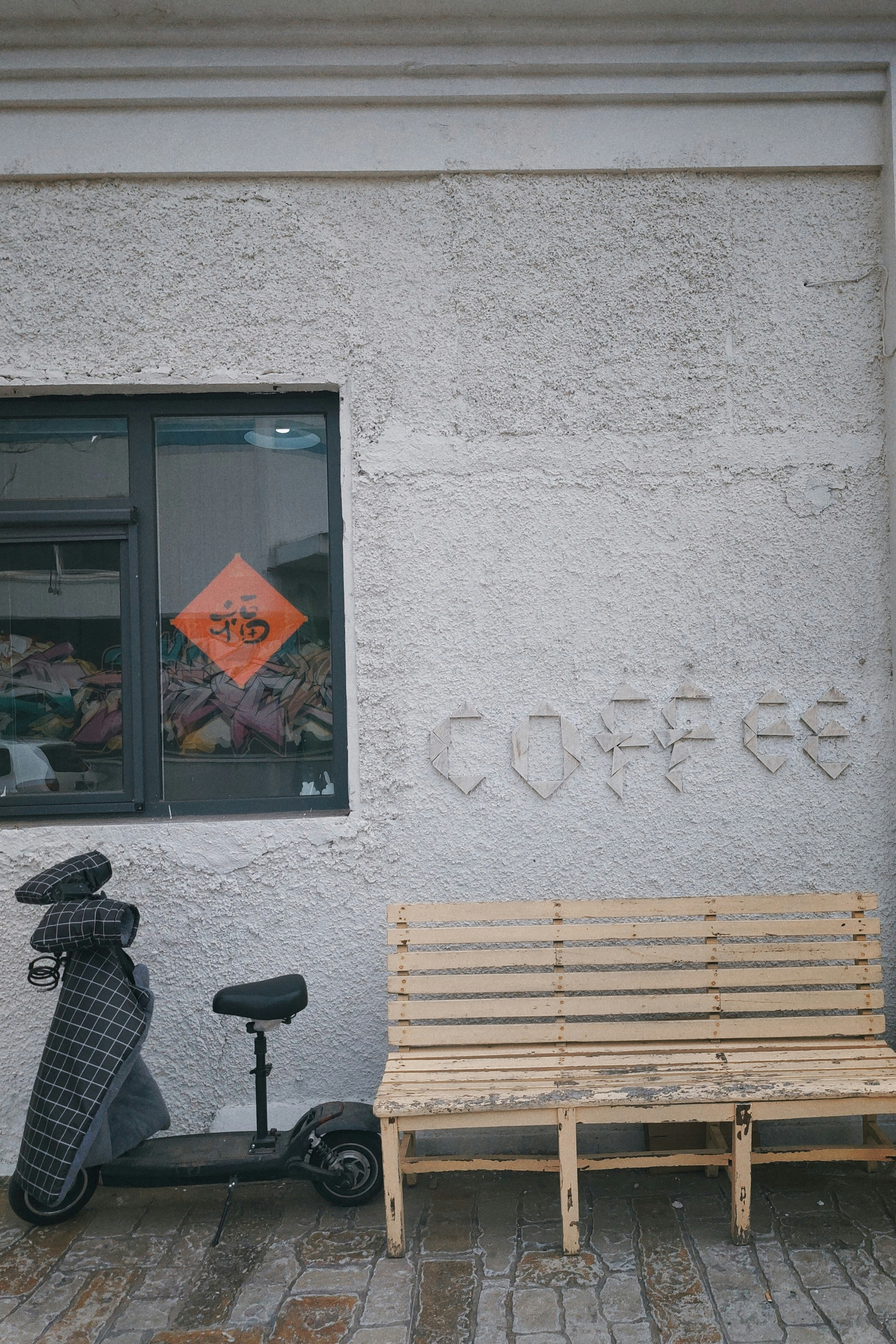 Storefront window features a bright orange fortune diamond against a textured wall. A wooden bench and a covered scooter rest on a cobblestone street.