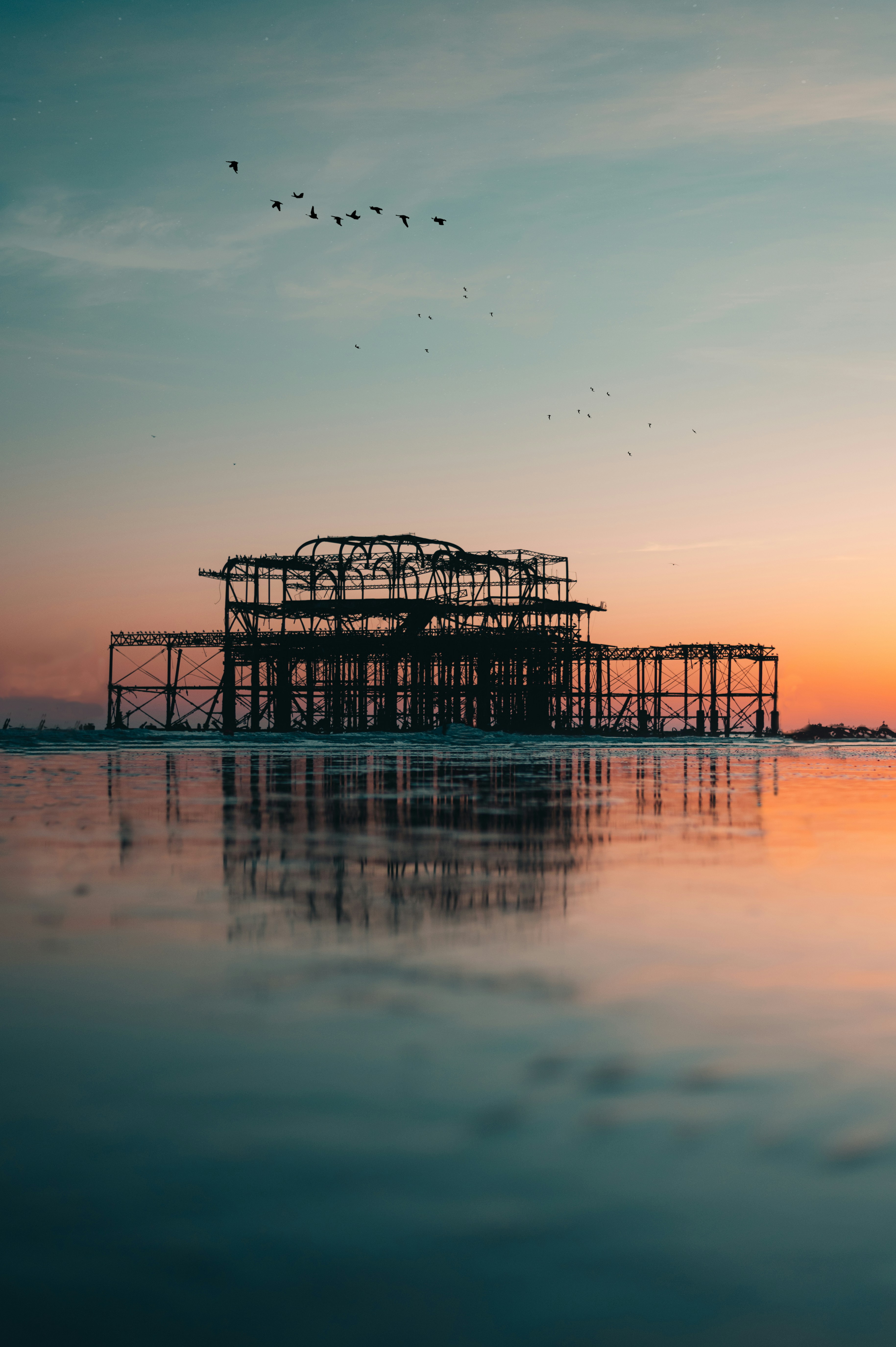 Silhouette of a derelict pier against a vibrant sunset sky, with birds flying overhead. Reflections shimmer in the water below.