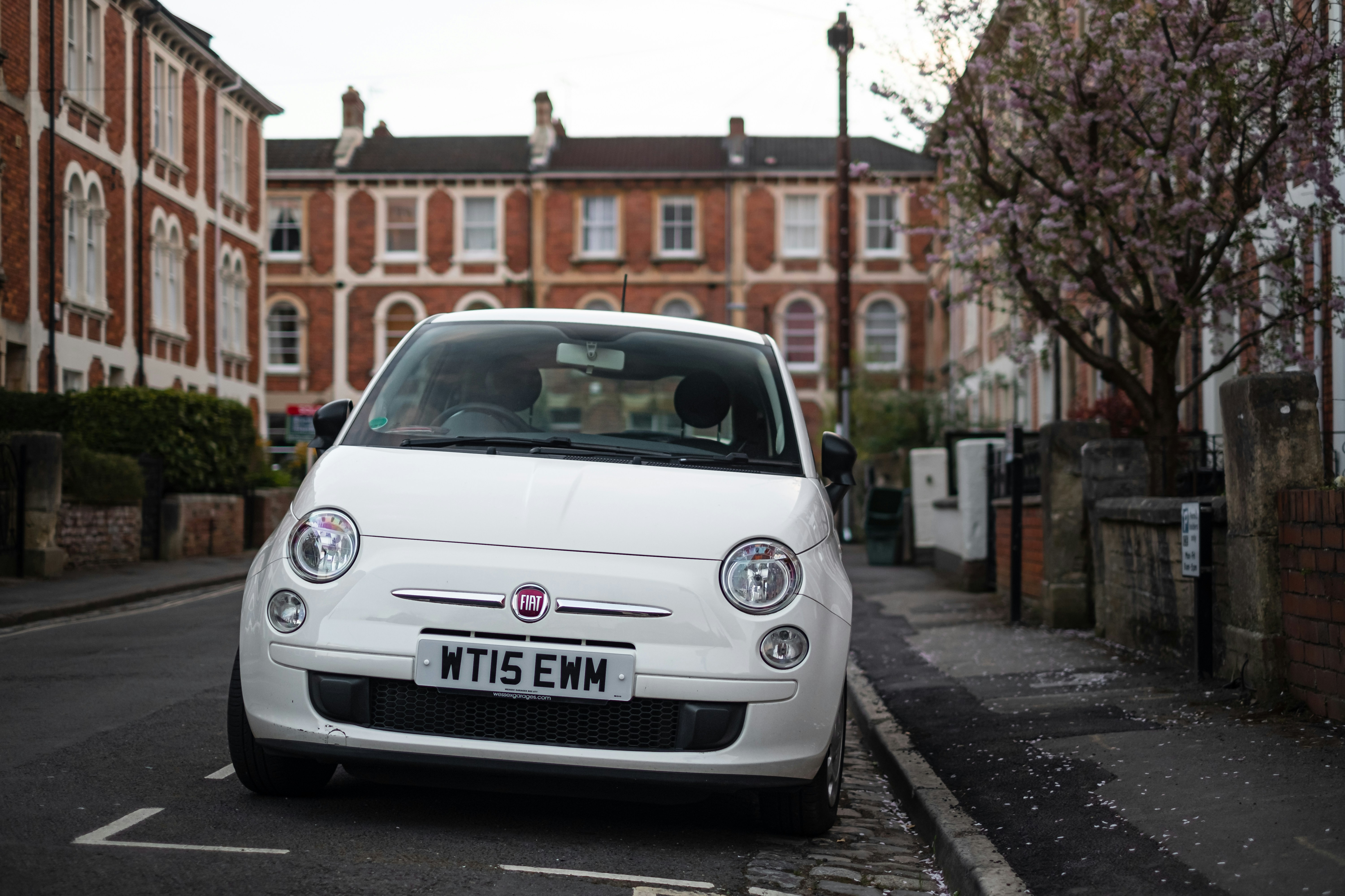 A white Fiat parked on a quaint street lined with brick buildings and flowering trees, showcasing urban life.