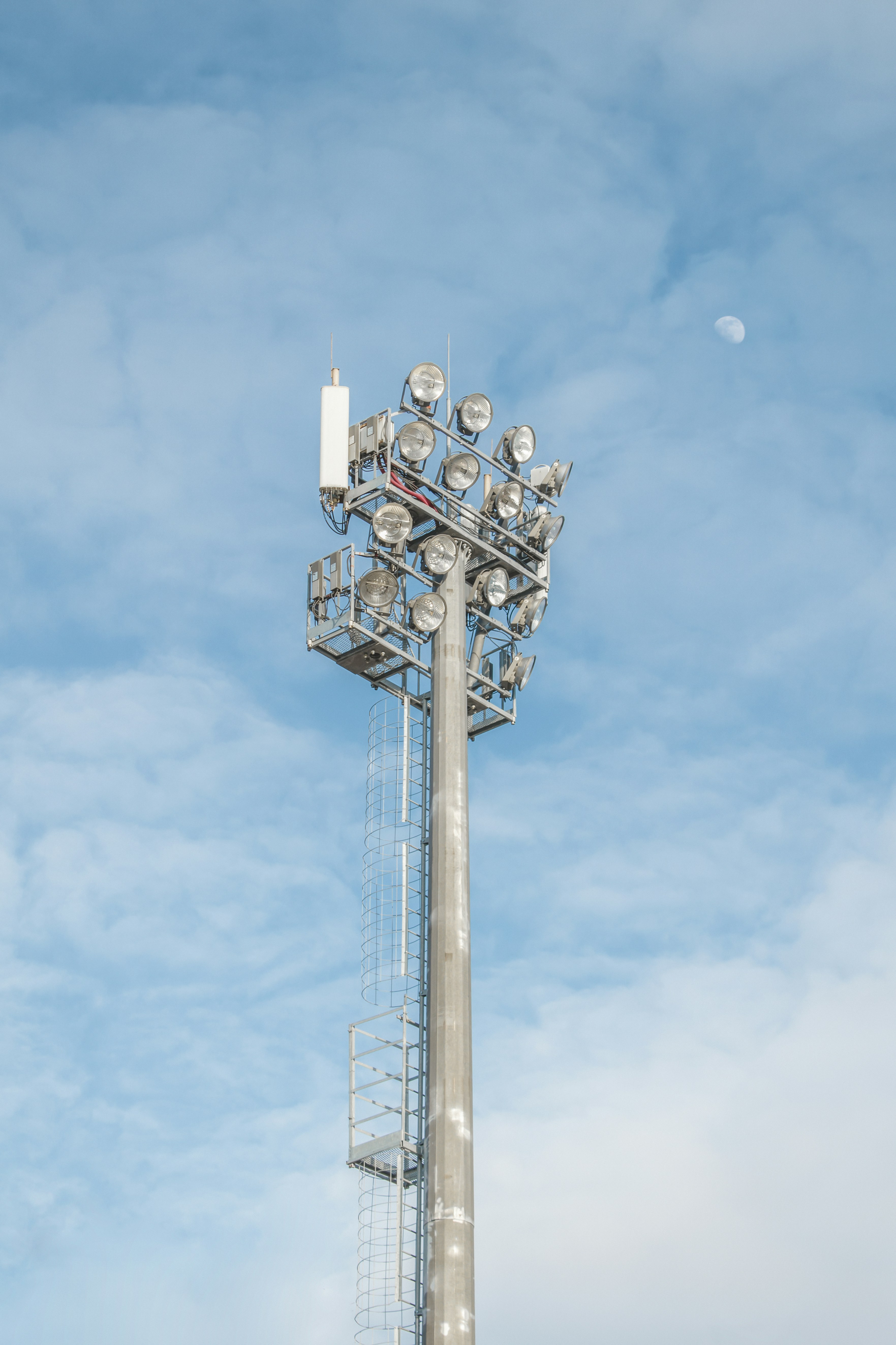Tall floodlight tower with multiple lamps against a backdrop of blue sky and a visible moon.
