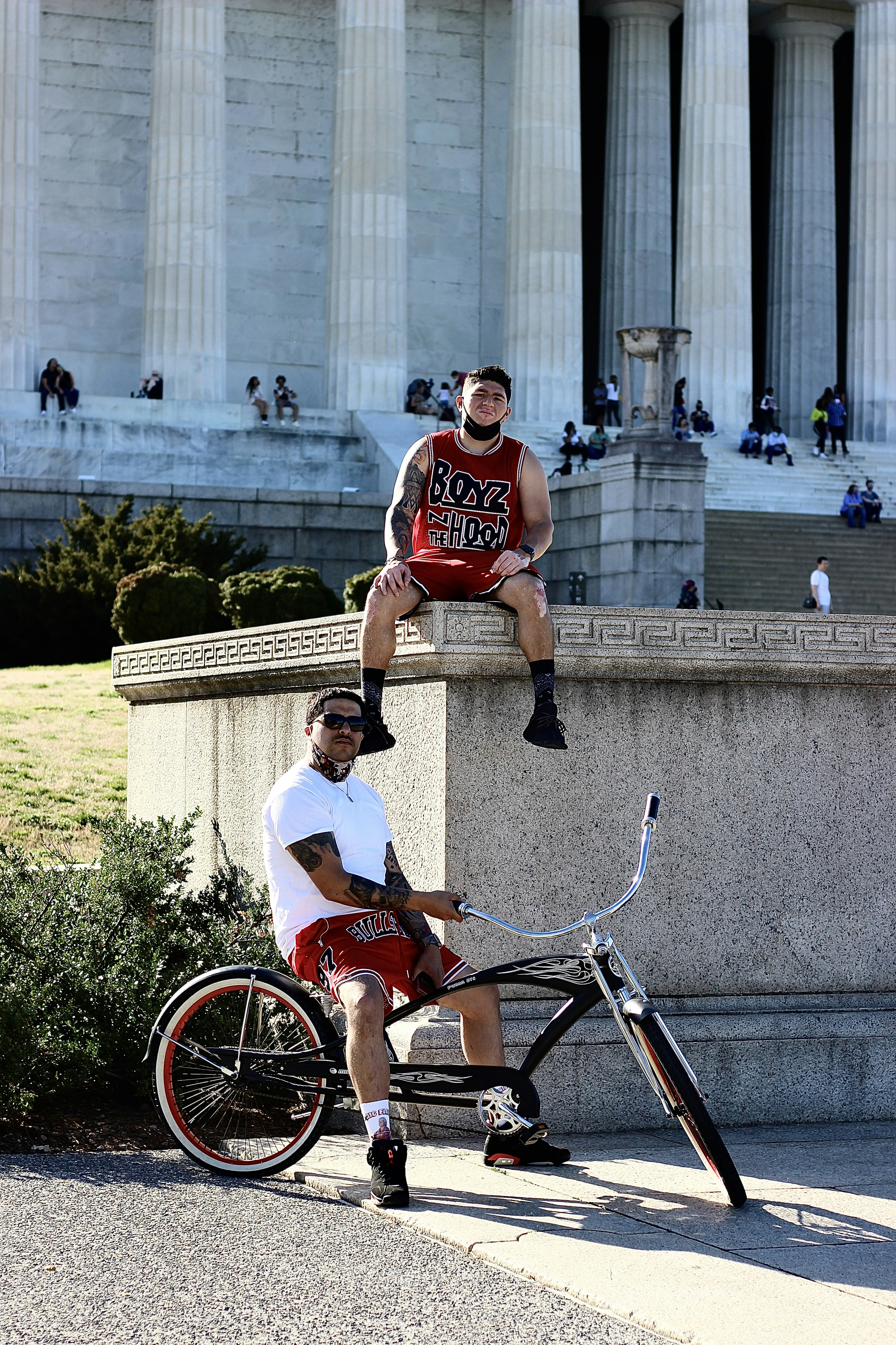 Two individuals in vibrant streetwear pose with a custom bicycle near the iconic Lincoln Memorial, showcasing a blend of culture and history.