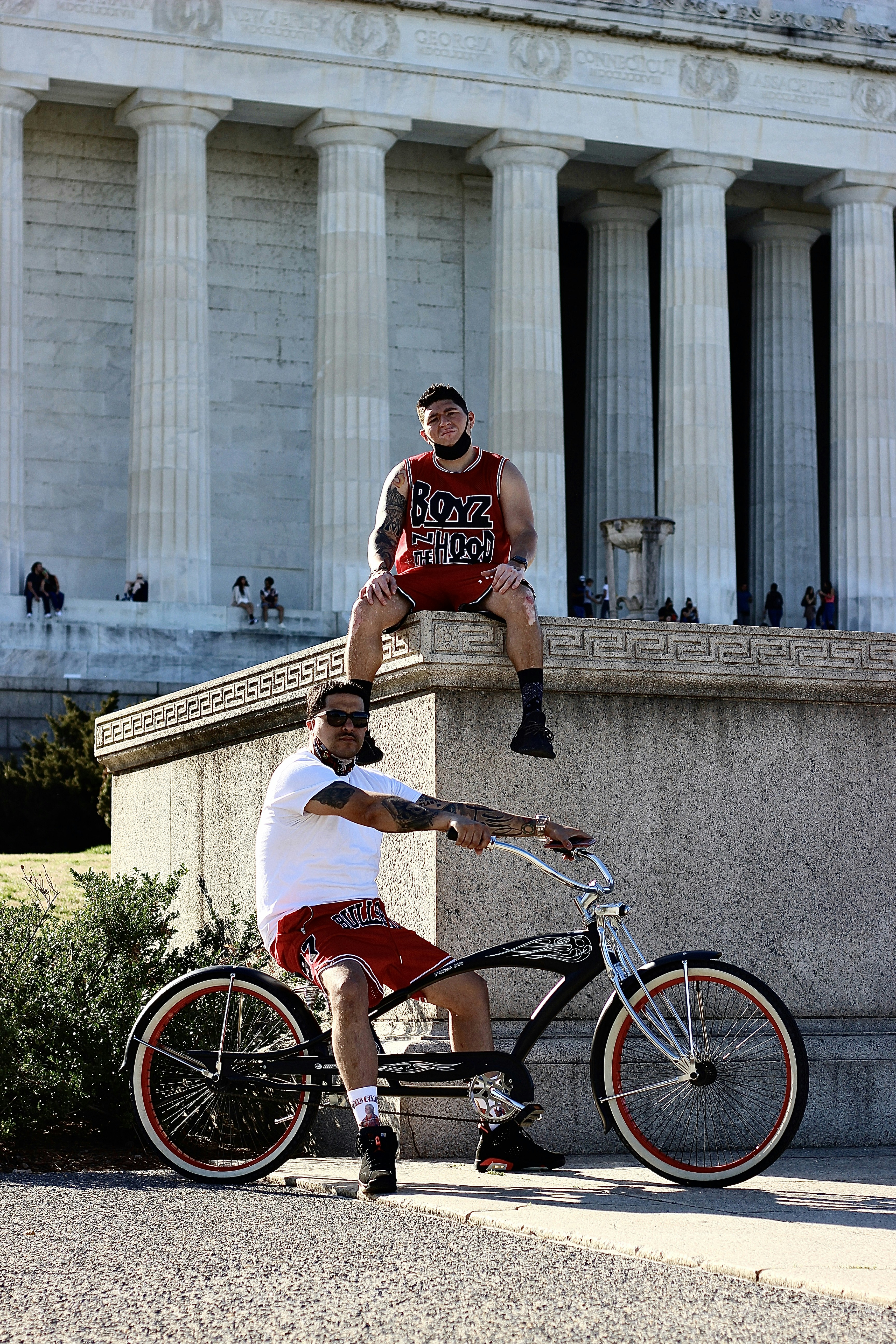 Two men in vibrant streetwear pose with a custom bicycle in front of the iconic Lincoln Memorial, showcasing a blend of culture and history.