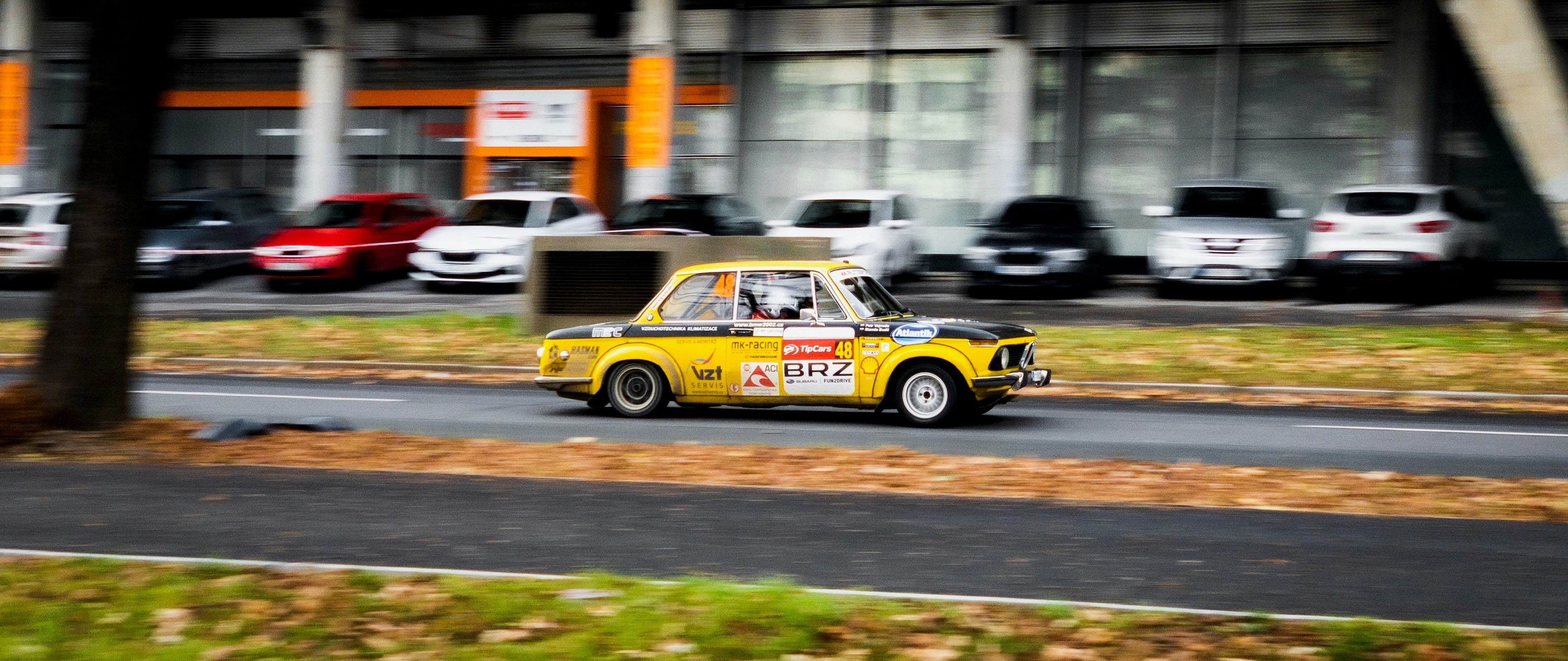A classic yellow car speeds down a city street, surrounded by modern vehicles and fallen leaves. The dynamic motion captures the essence of automotive nostalgia.