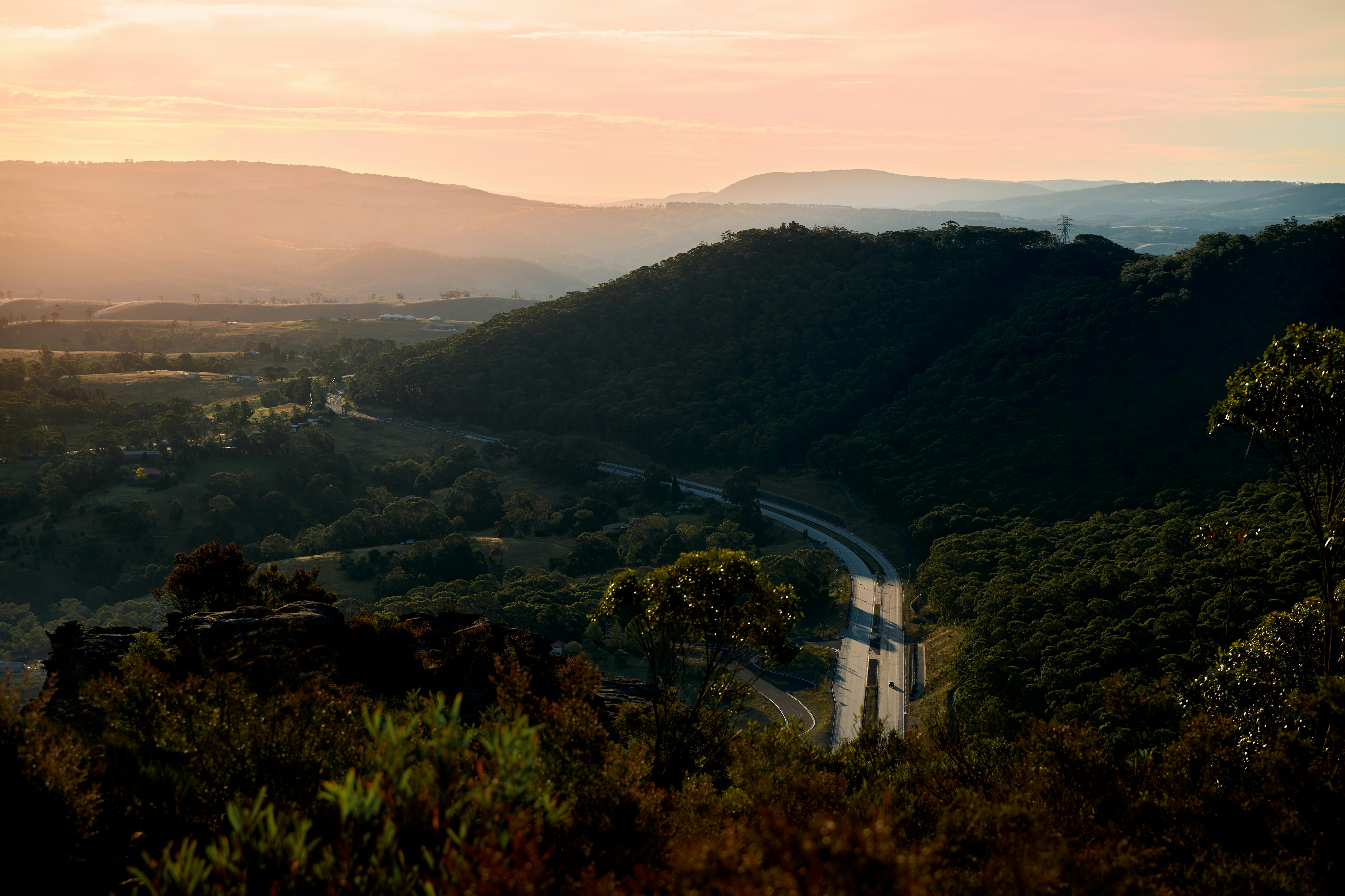 Curving road meanders through lush hills under a soft sunset glow, highlighting the serene landscape. The scene captures the transition from day to night.