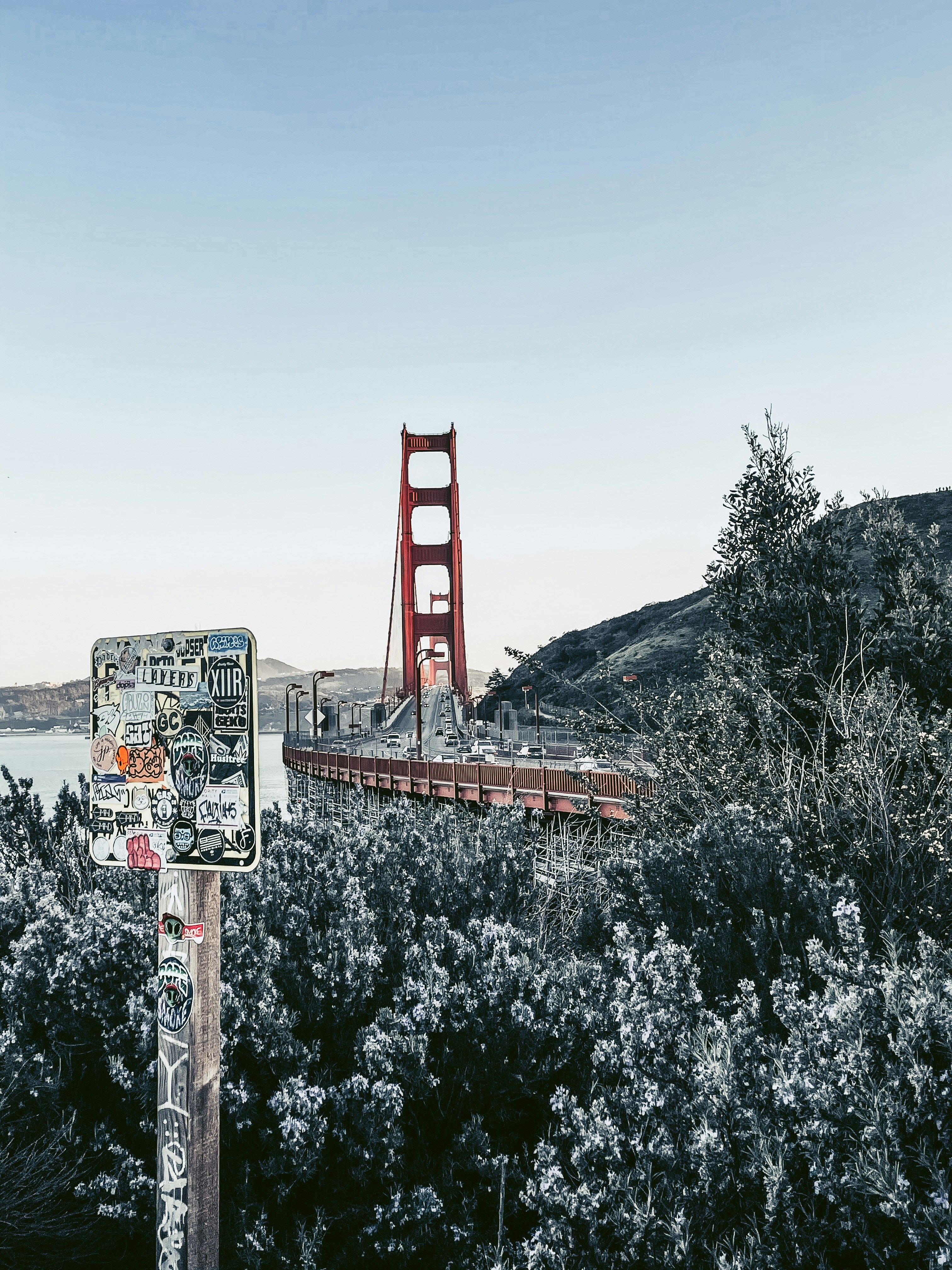 A vibrant sticker-covered sign stands in the foreground, framing the iconic Golden Gate Bridge in the background, surrounded by lush greenery.