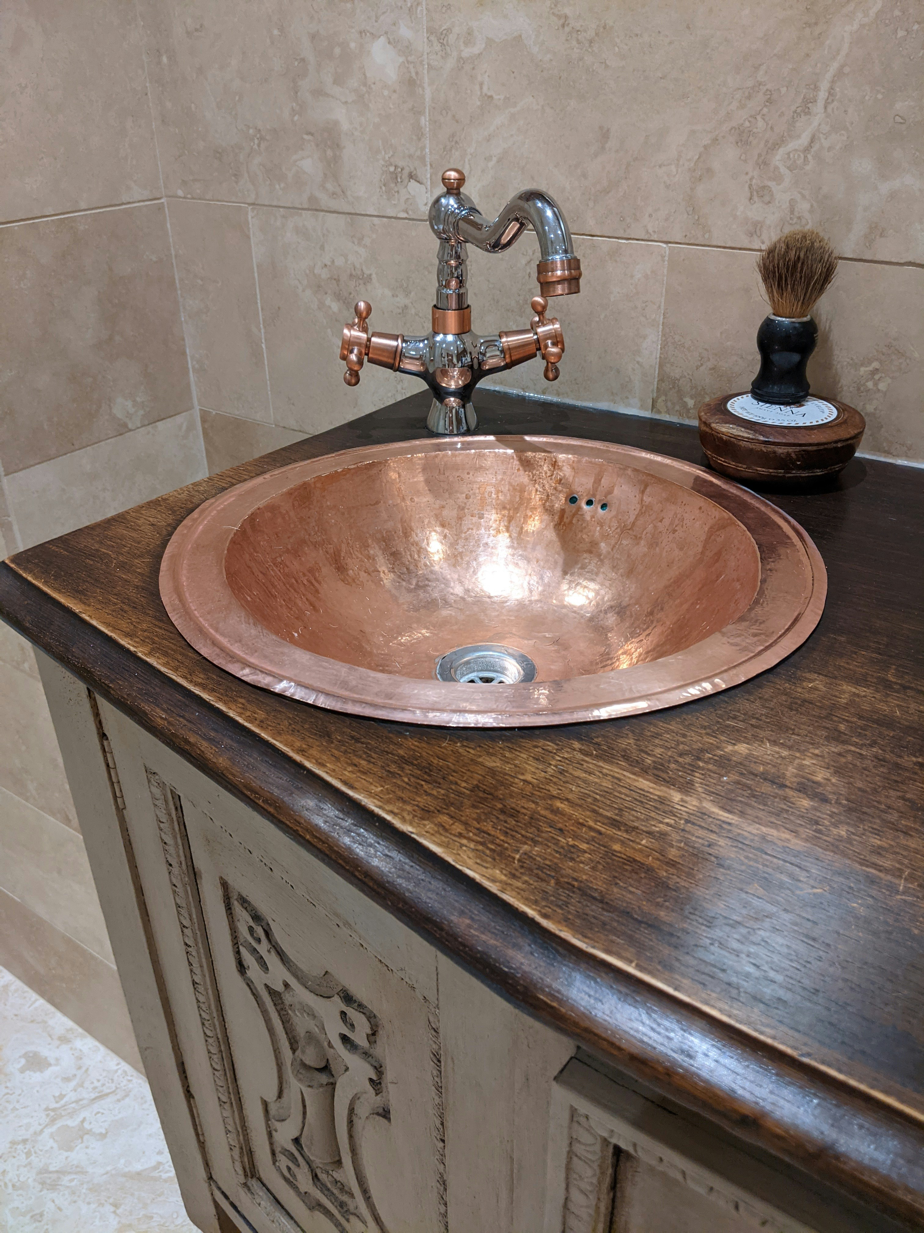Copper basin set into a dark wooden vanity, framed by beige tiled walls with a vintage cross-handle faucet.