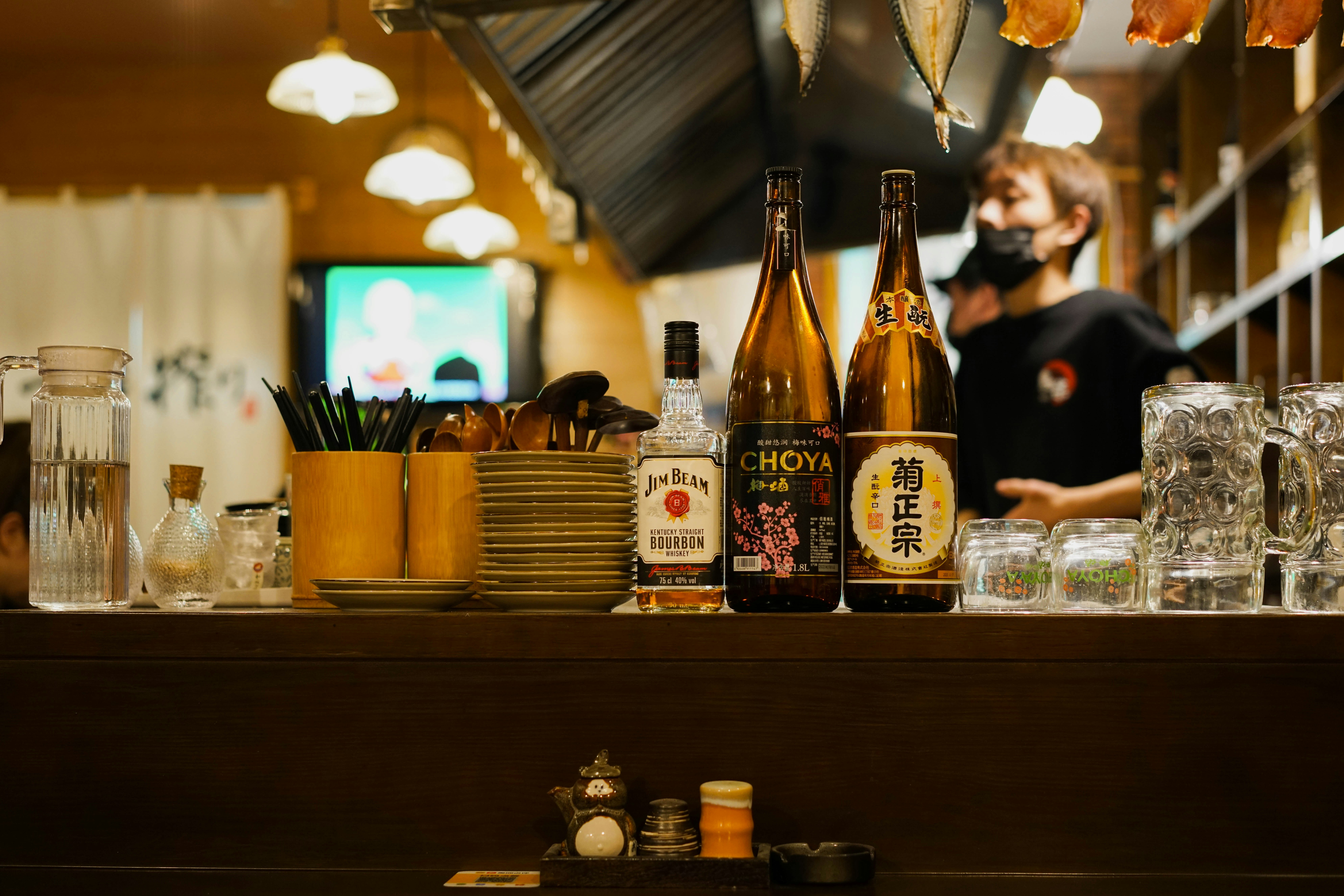 Bottles and glasses line a warmly lit bar counter, with a bartender in the background.