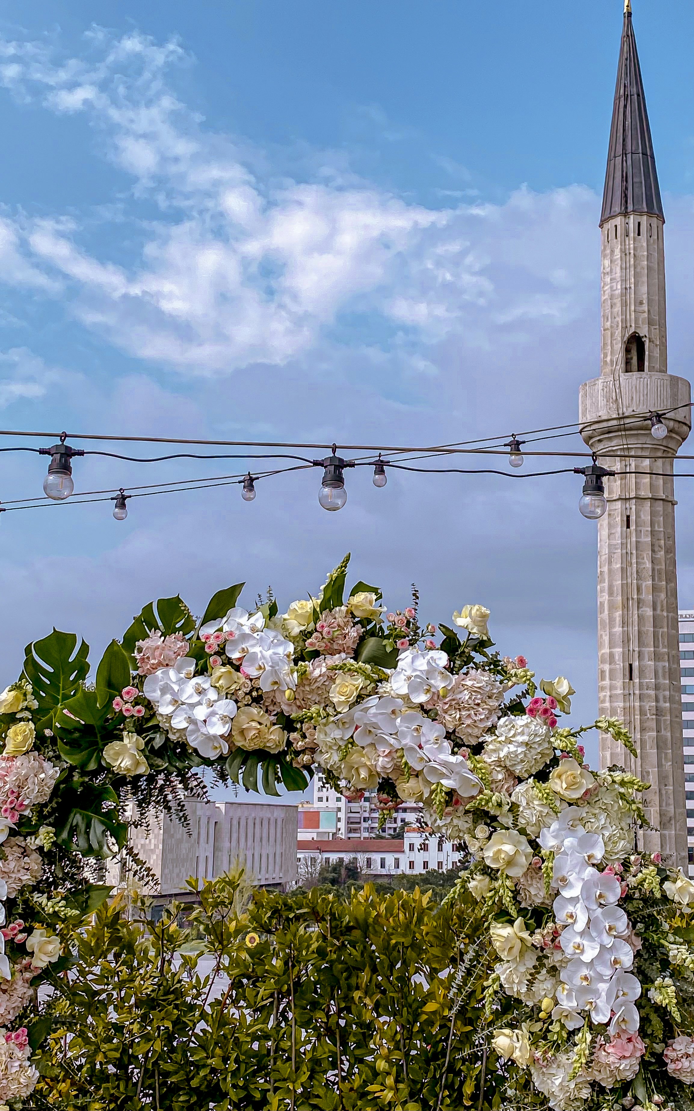 A floral arch adorned with white orchids and greenery frames a city skyline, with a tall minaret visible in the background under a blue sky.