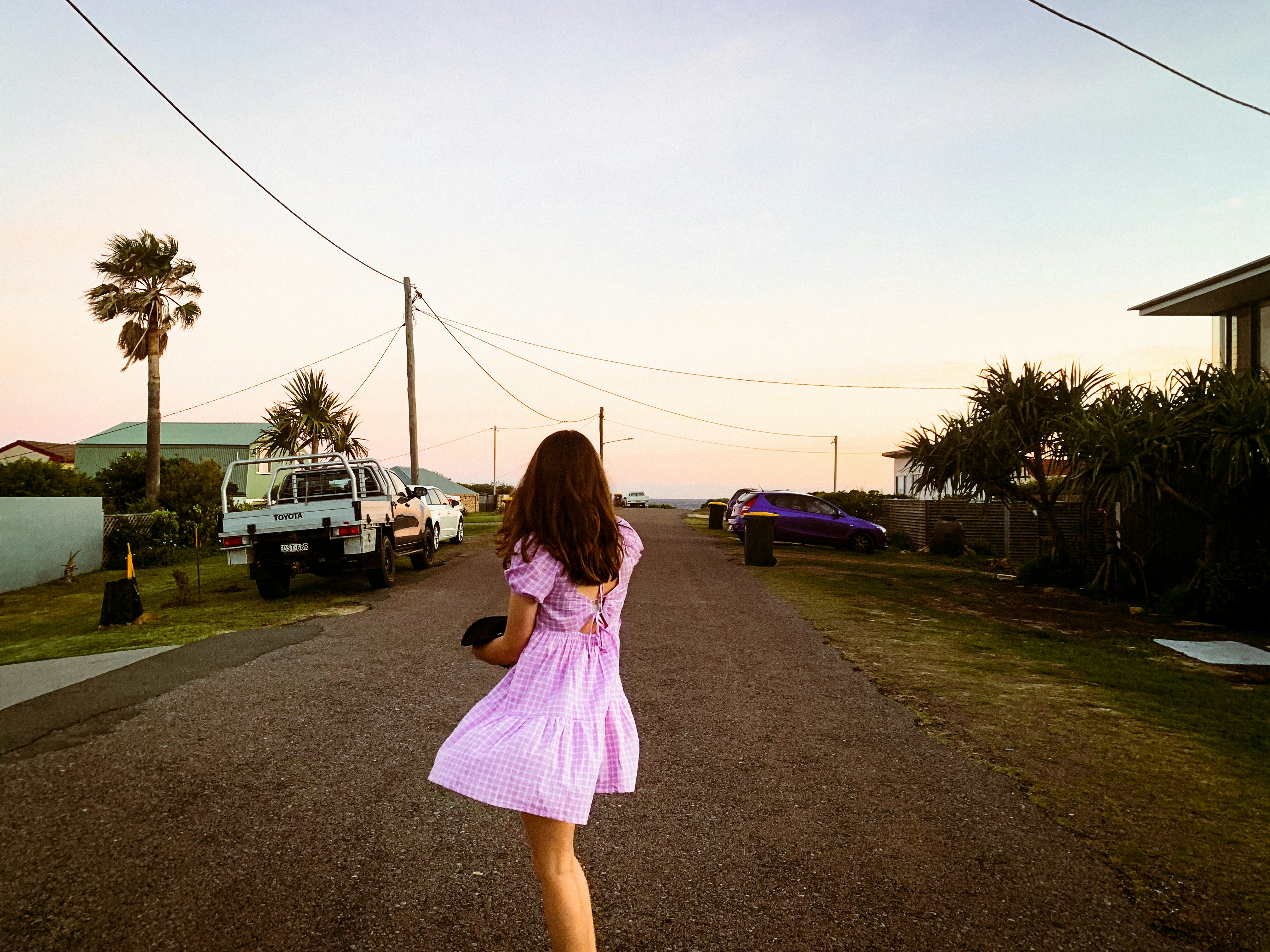Woman in a purple dress walking down a suburban street at sunset with palm trees and parked cars.