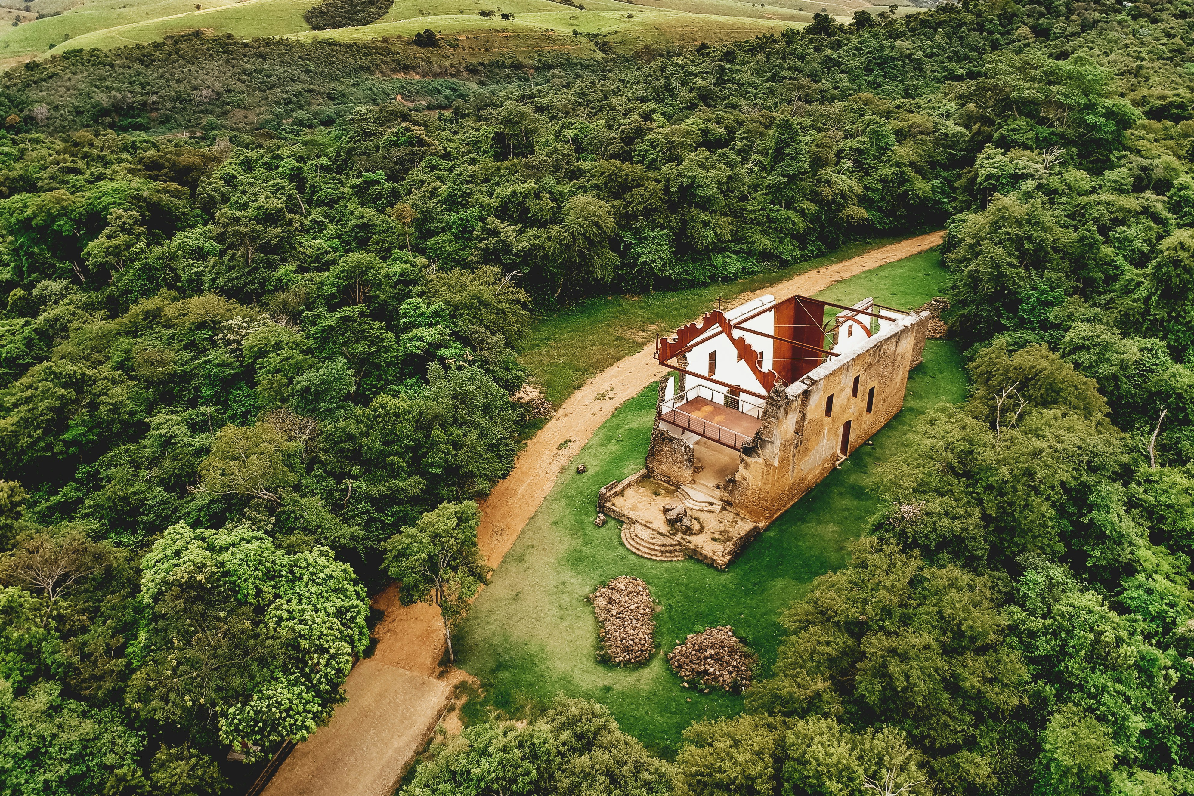 Aerial view of a partially ruined building surrounded by dense green forest and a winding dirt path.