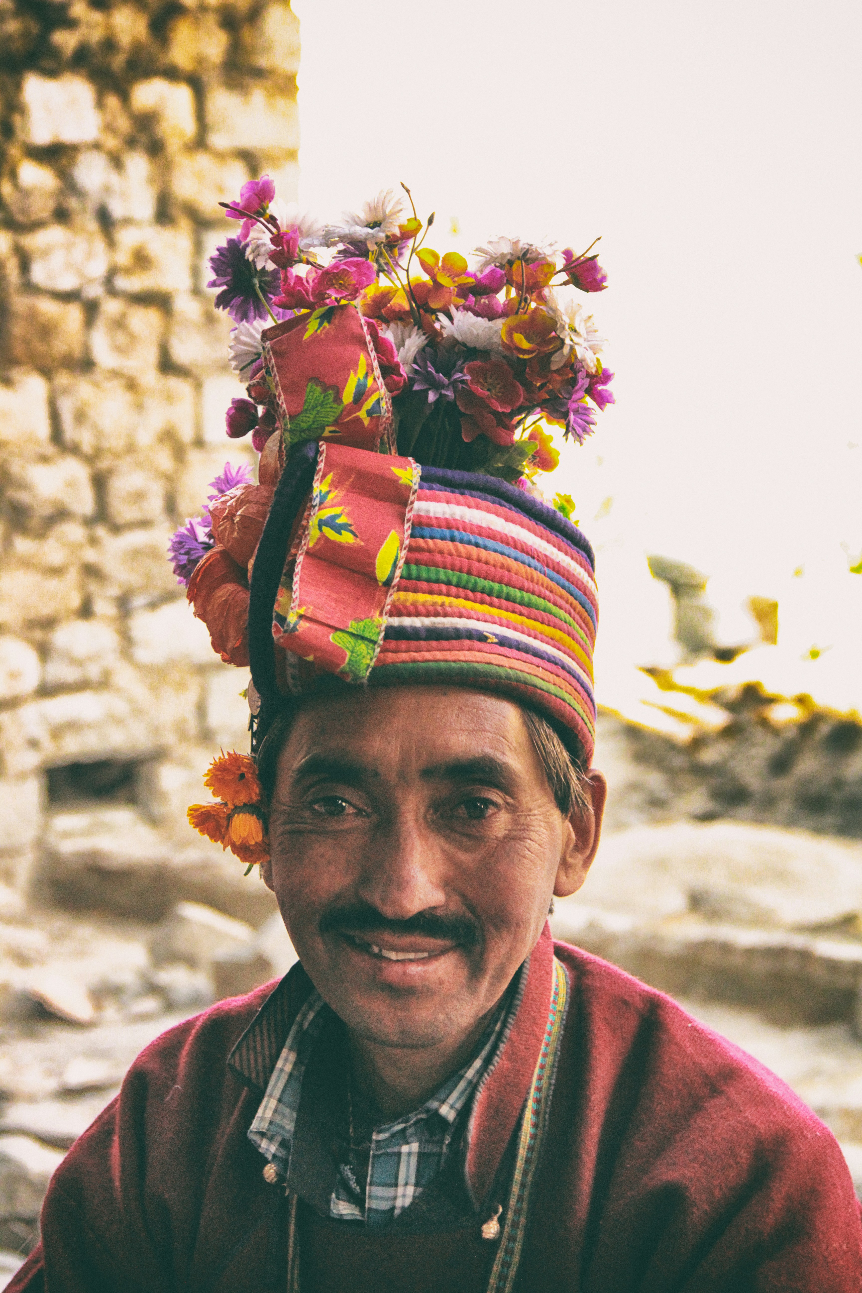 A smiling man adorned with a vibrant floral headdress, showcasing traditional craftsmanship against a rustic stone backdrop.