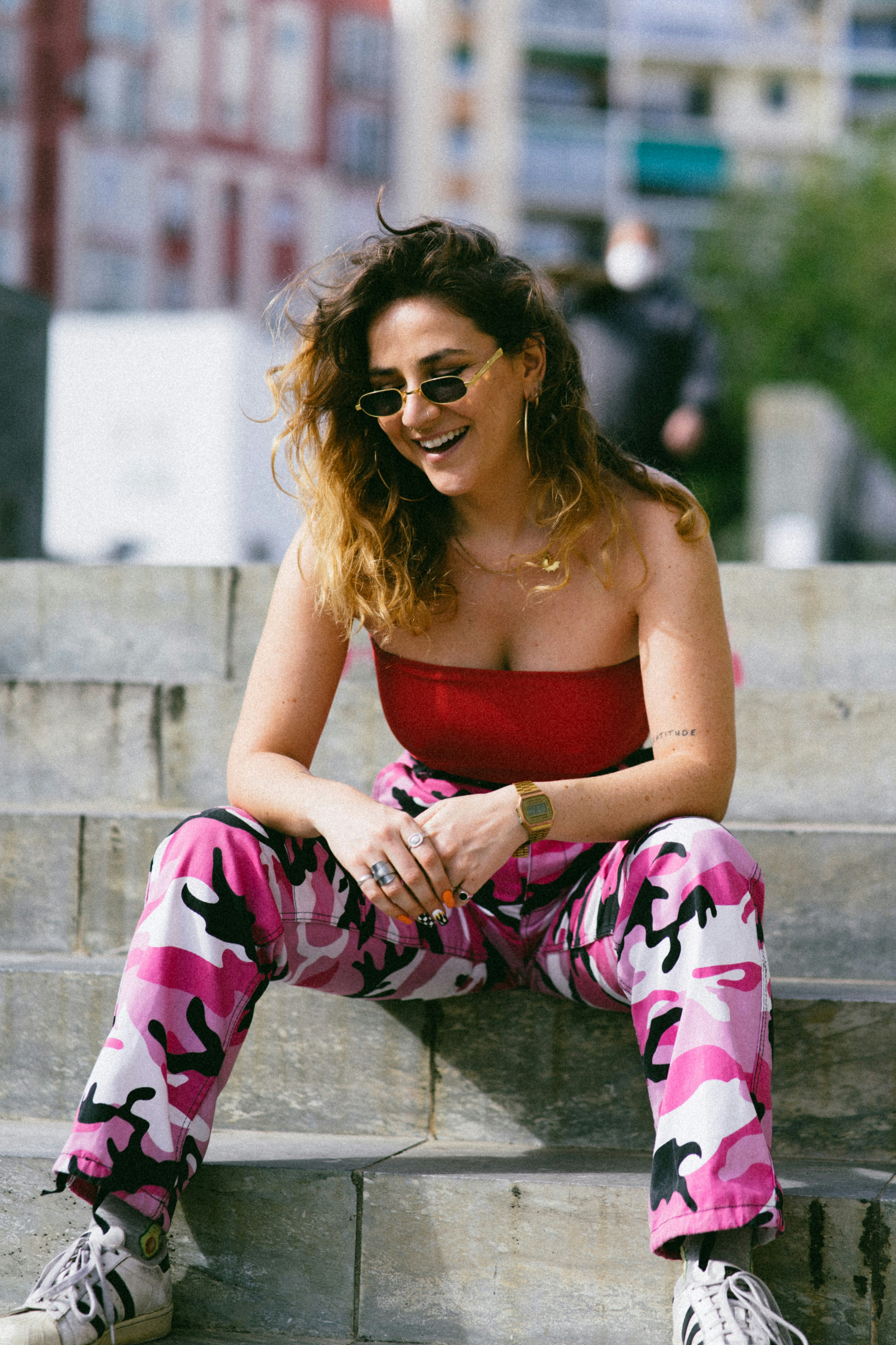 Woman laughing while seated on stone steps, wearing a red top and pink camouflage pants. Sunglasses add a stylish flair.