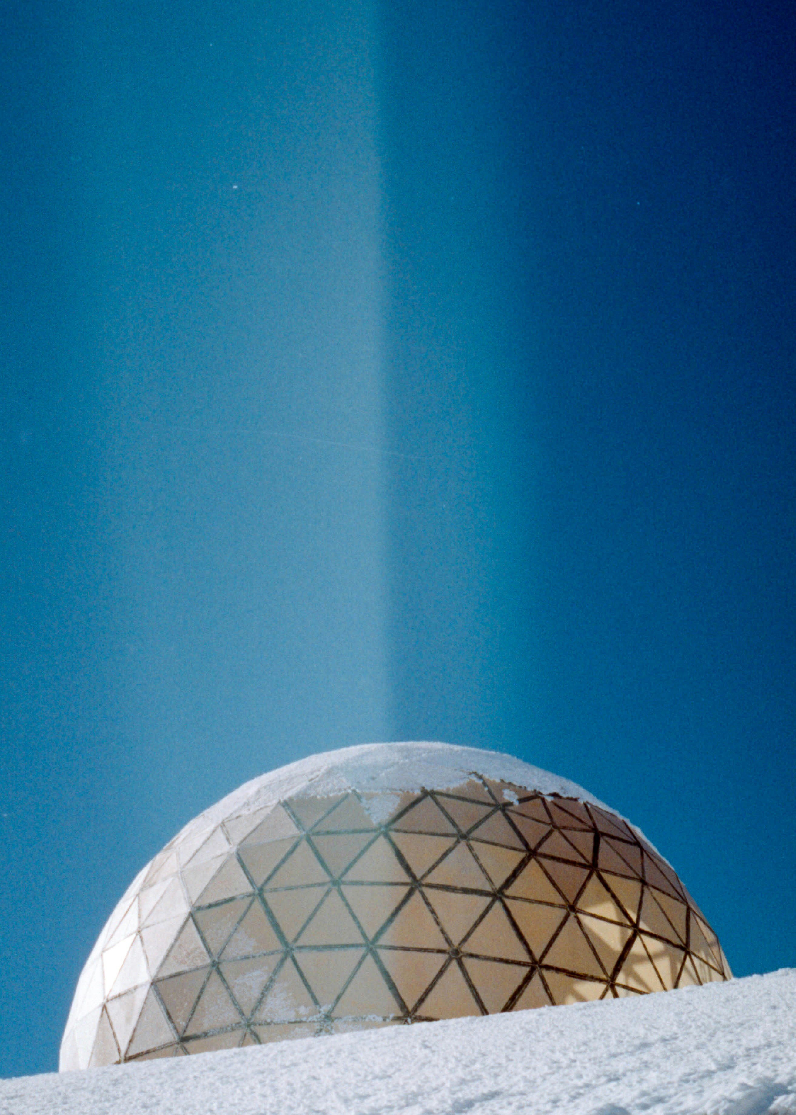 Geodesic dome partially covered in snow against a clear blue sky, with a subtle light beam illuminating the scene.