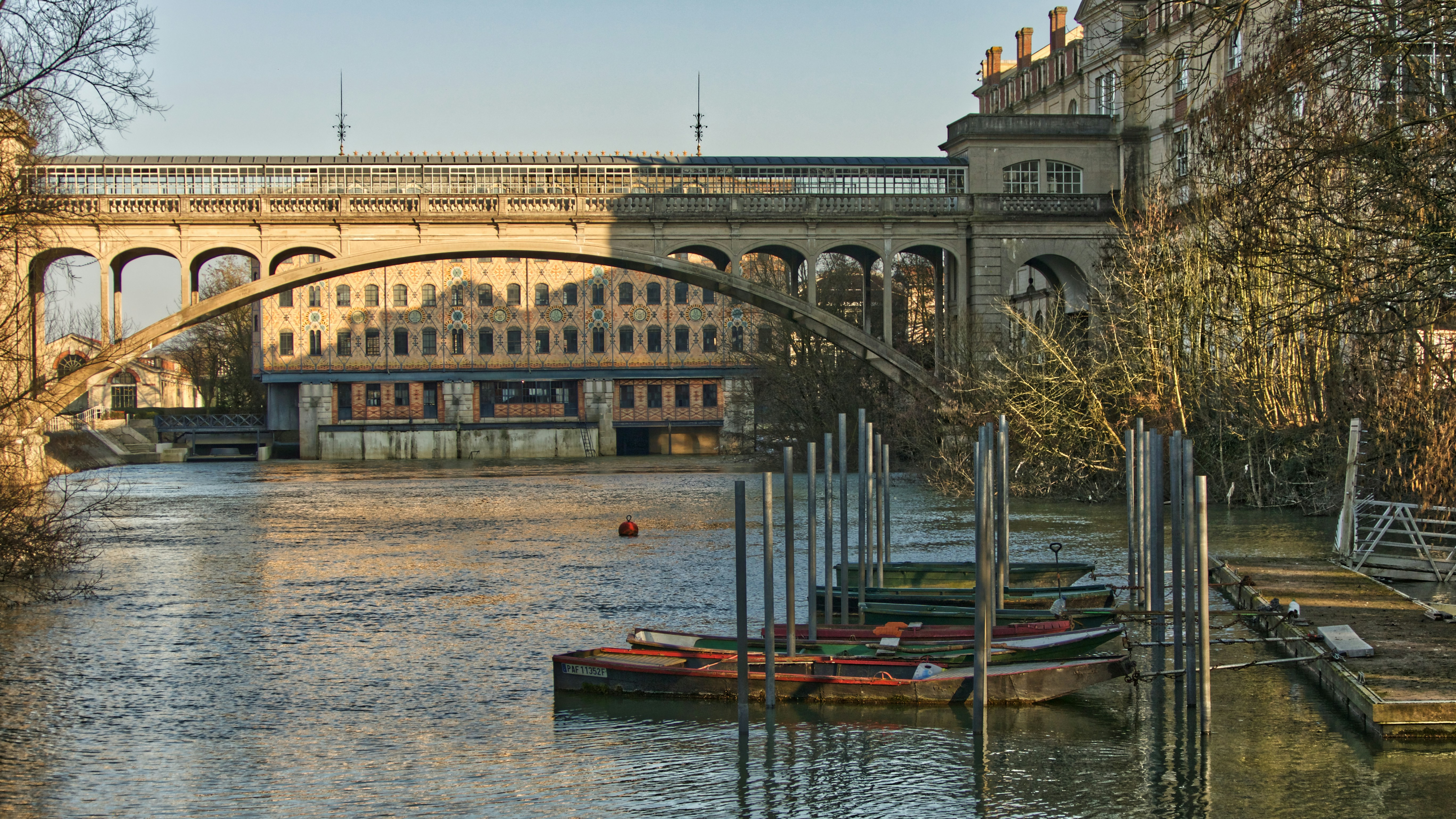 Historic factory building by a river, framed by an arched bridge and winter trees.