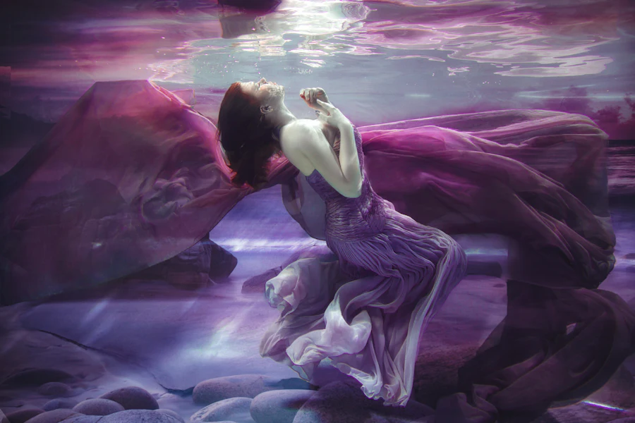 A woman poses underwater in a flowing purple gown, surrounded by rippling light and rocks. The scene has a dreamy, ethereal look with fabric drifting around her.