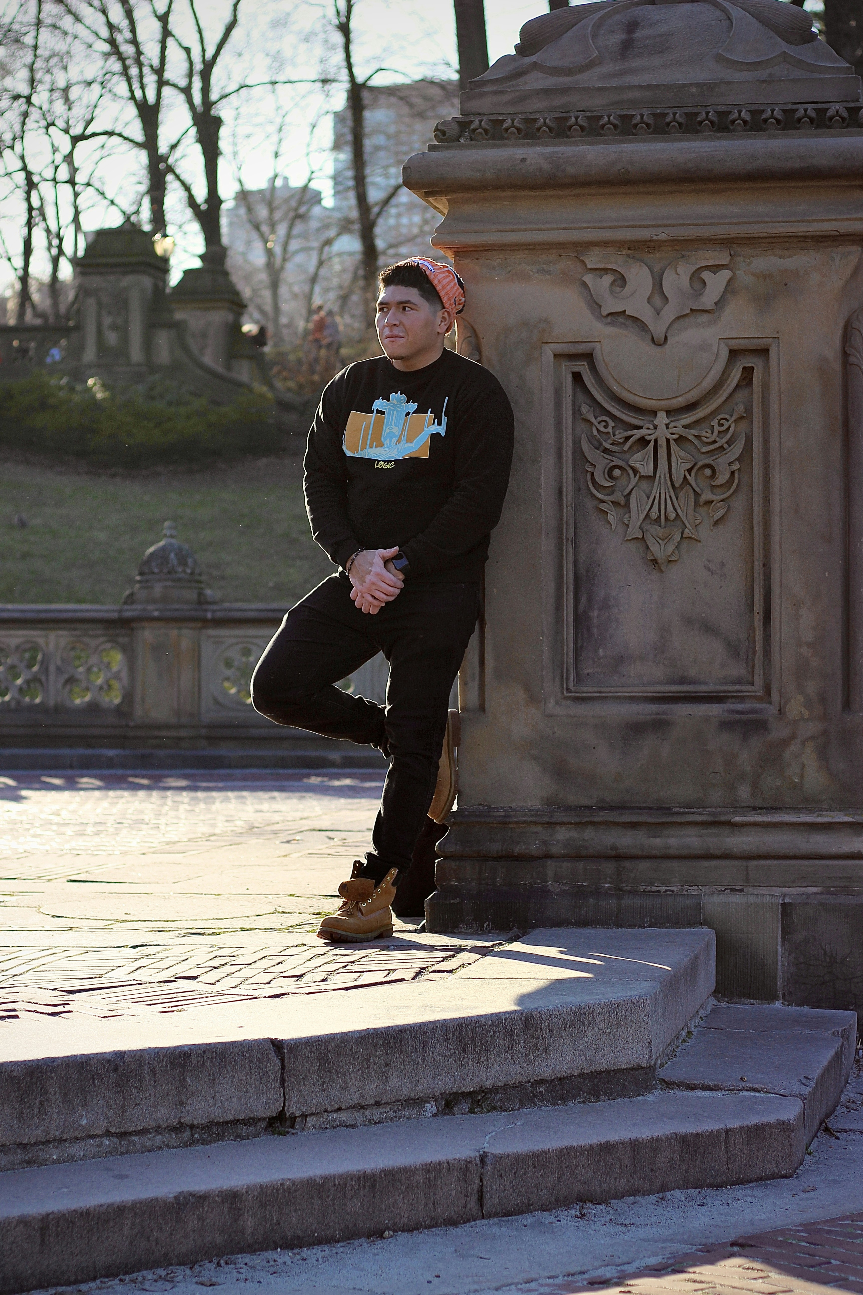 Individual leaning against an ornate stone structure in a sunlit urban park, exuding a sense of calm amidst the bustling backdrop.
