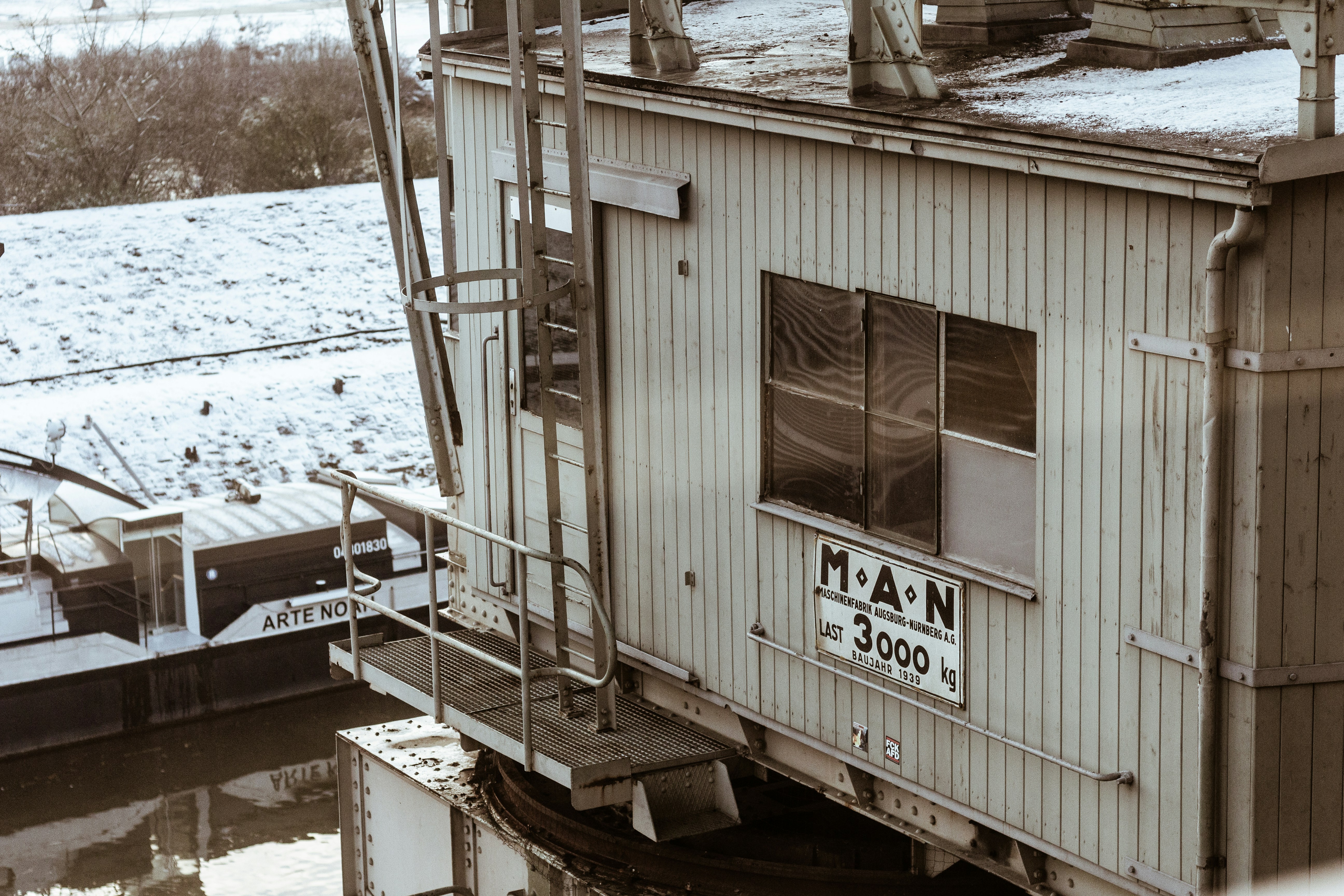 Snow-dusted industrial building beside a river, featuring a faded sign and large windows.