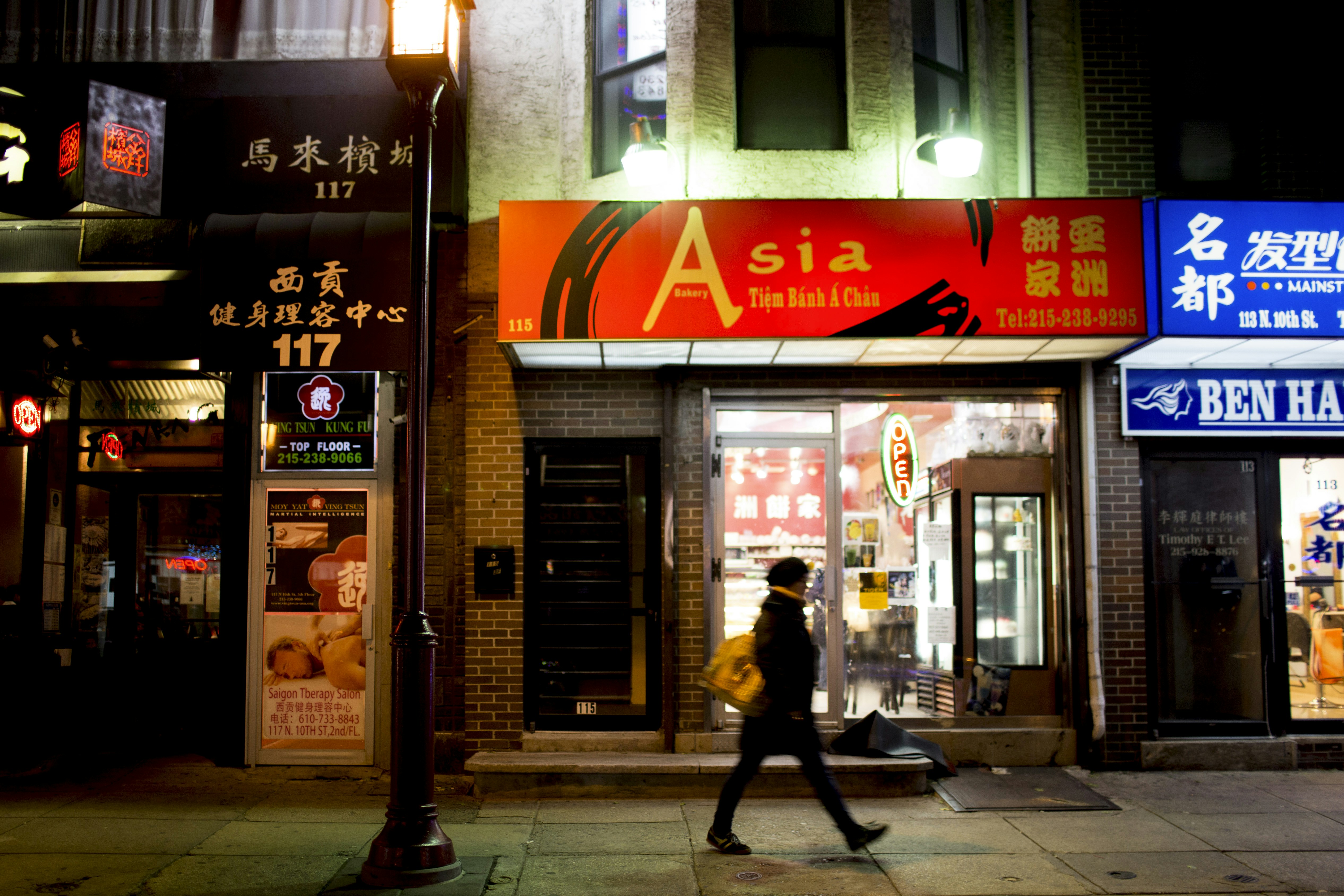 Person in black jacket walking past illuminated storefronts at night.