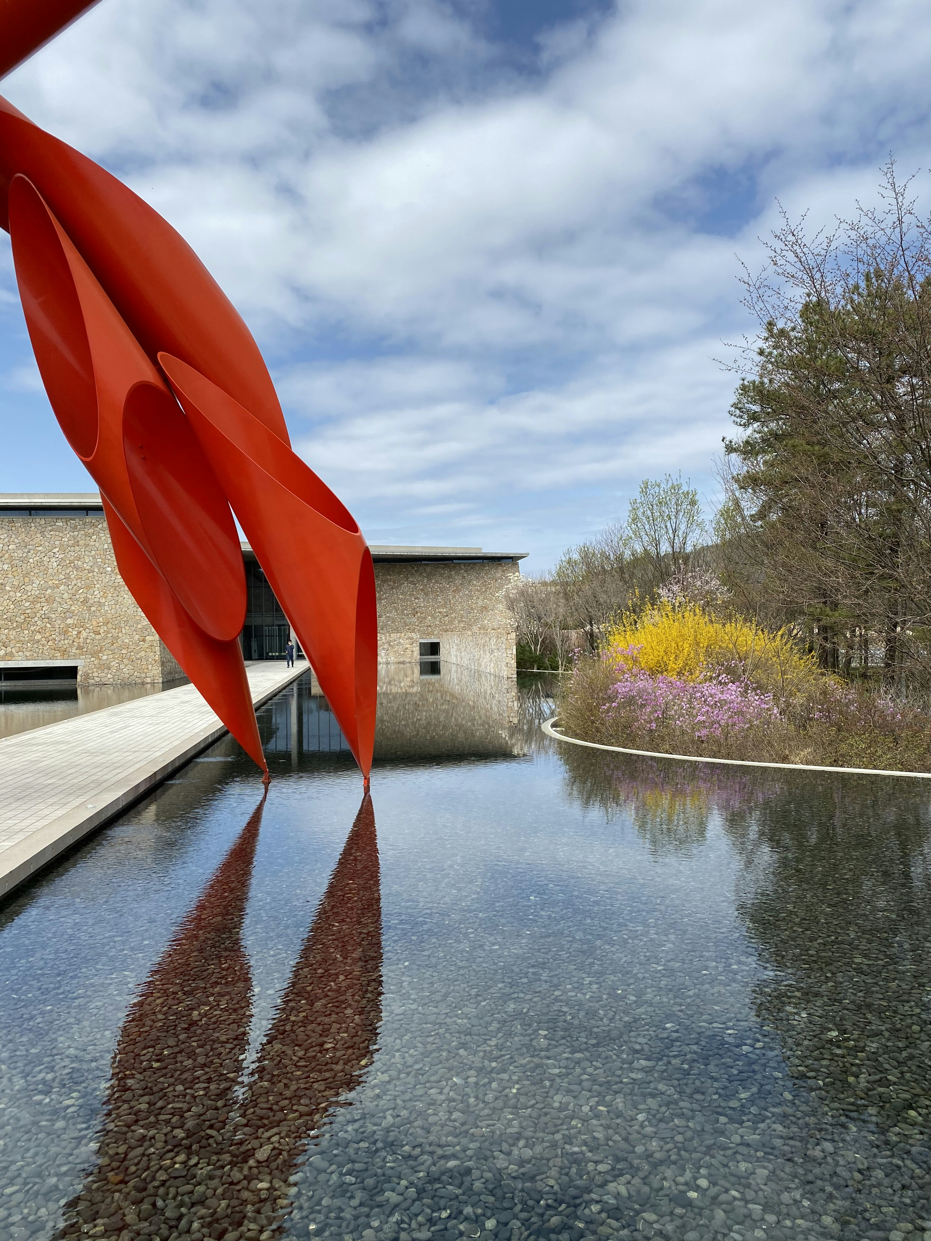 A striking red sculpture rises above a tranquil reflecting pool, surrounded by lush greenery and blooming flowers, showcasing the synergy between art and nature.