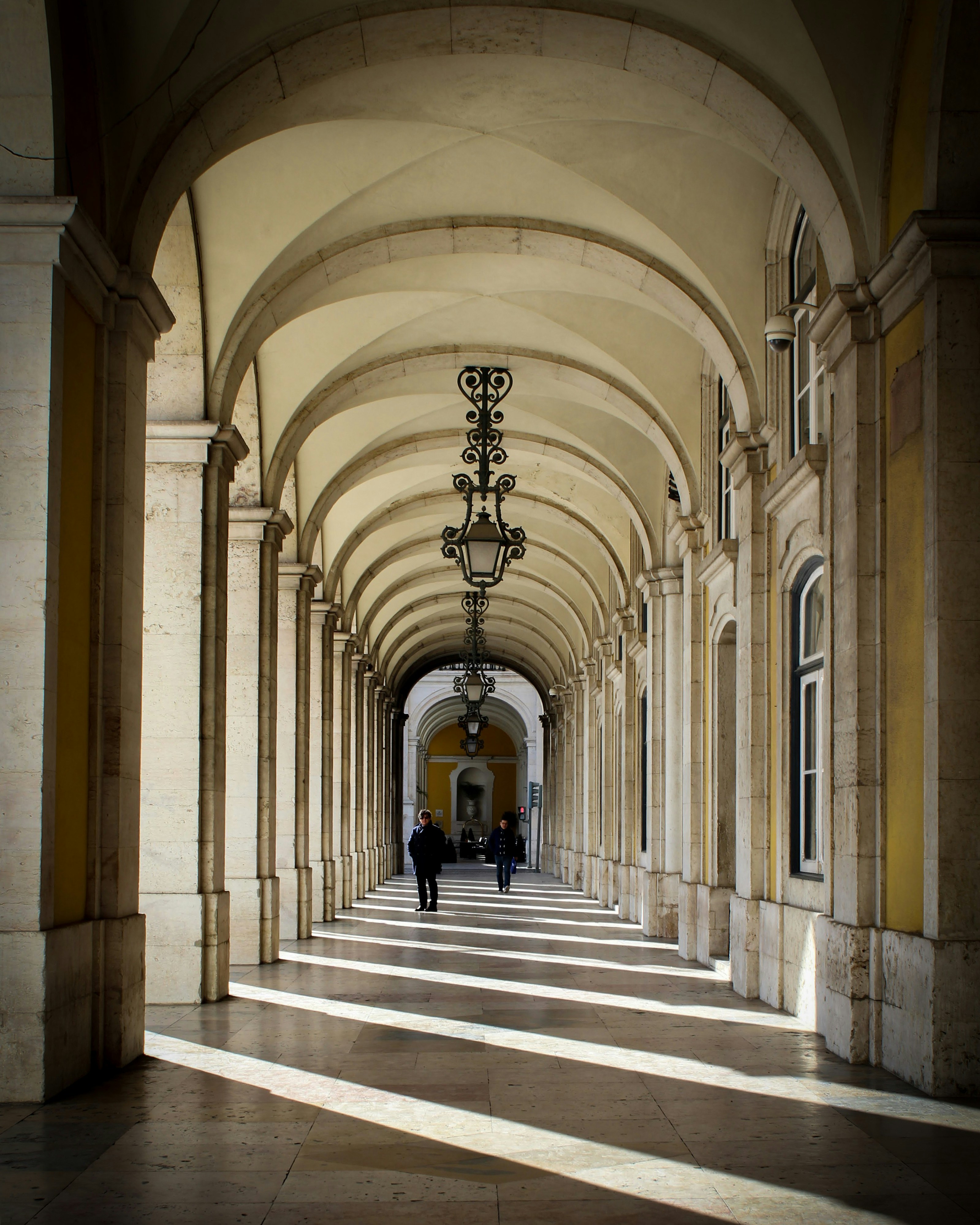 Elegant corridor featuring arched ceilings and intricate chandeliers, with two figures walking amidst long shadows. 
