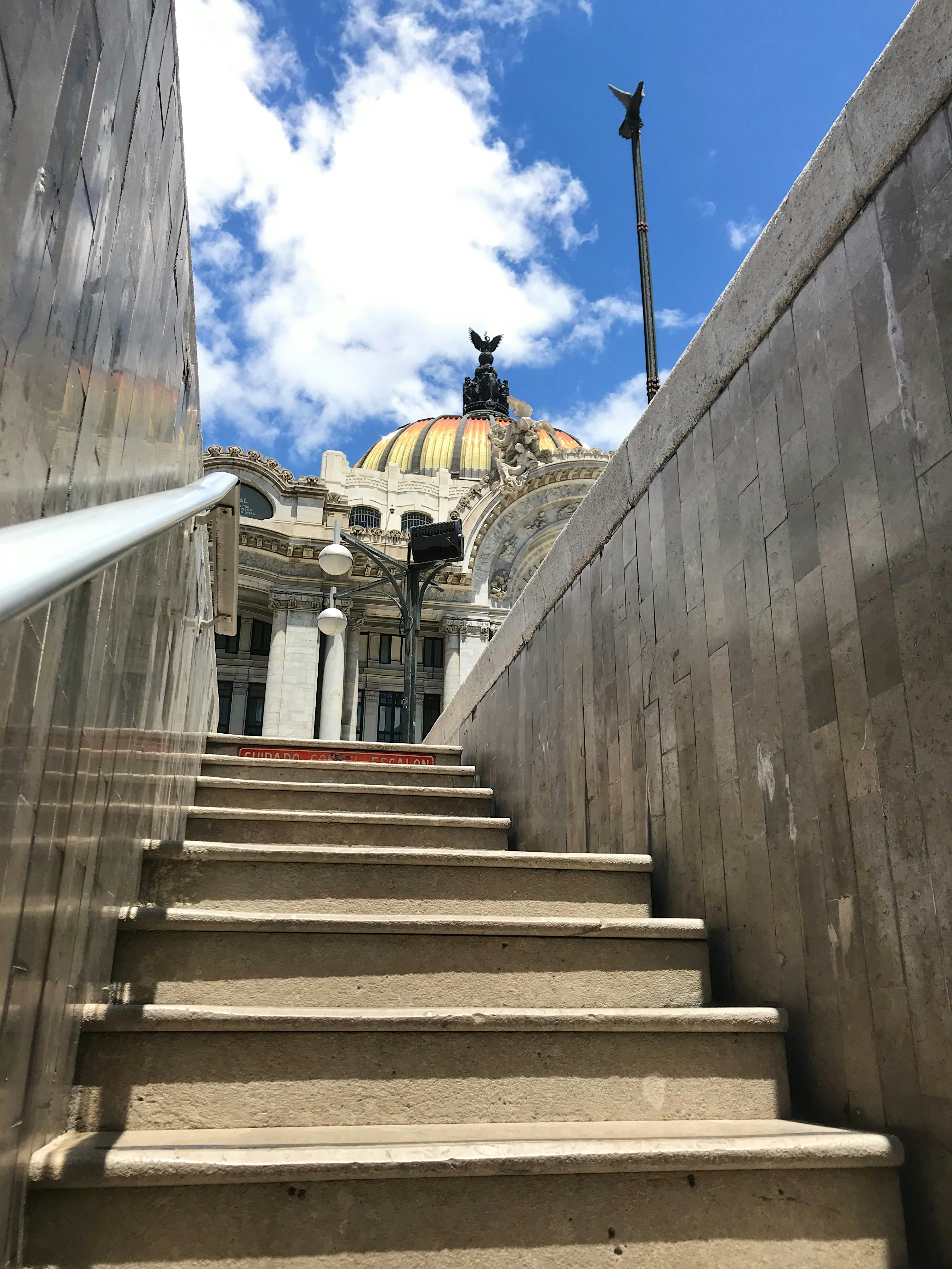 Stairs leading upward towards a grand building adorned with a colorful dome and intricate sculptures against a bright blue sky.