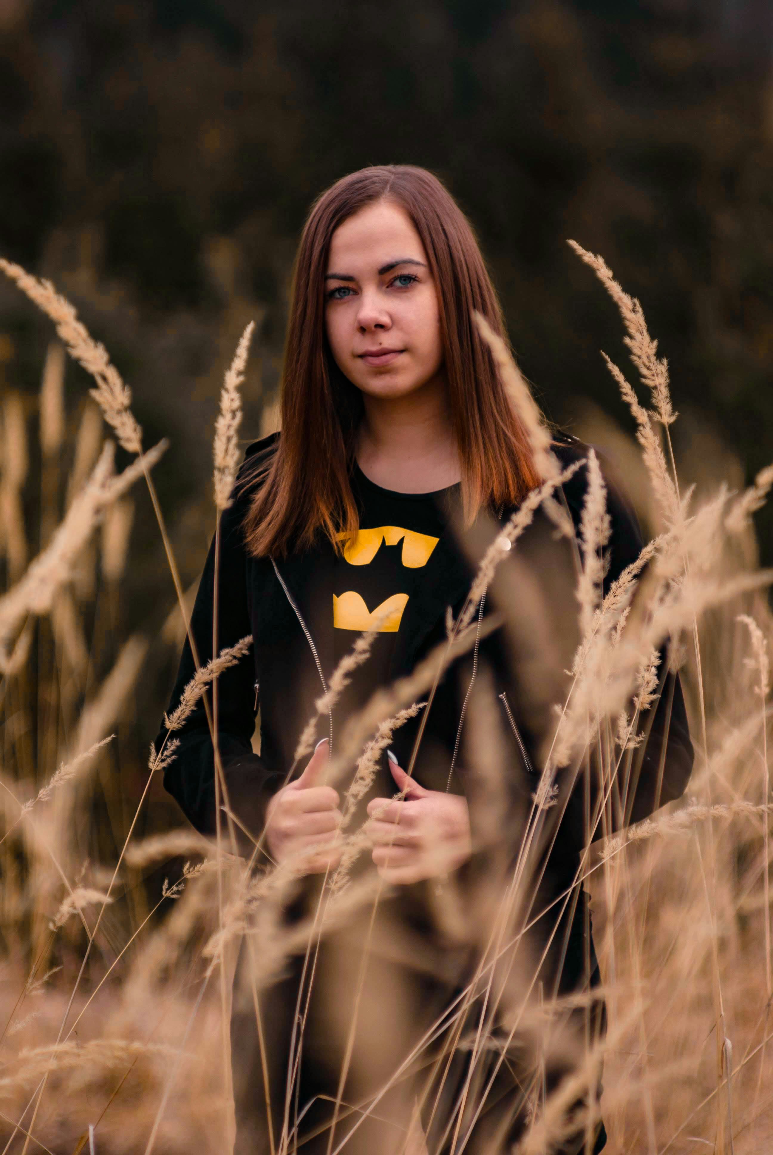 Portrait of a young woman in a black shirt with a yellow Batman emblem, standing among tall, golden reeds in a softly blurred field.