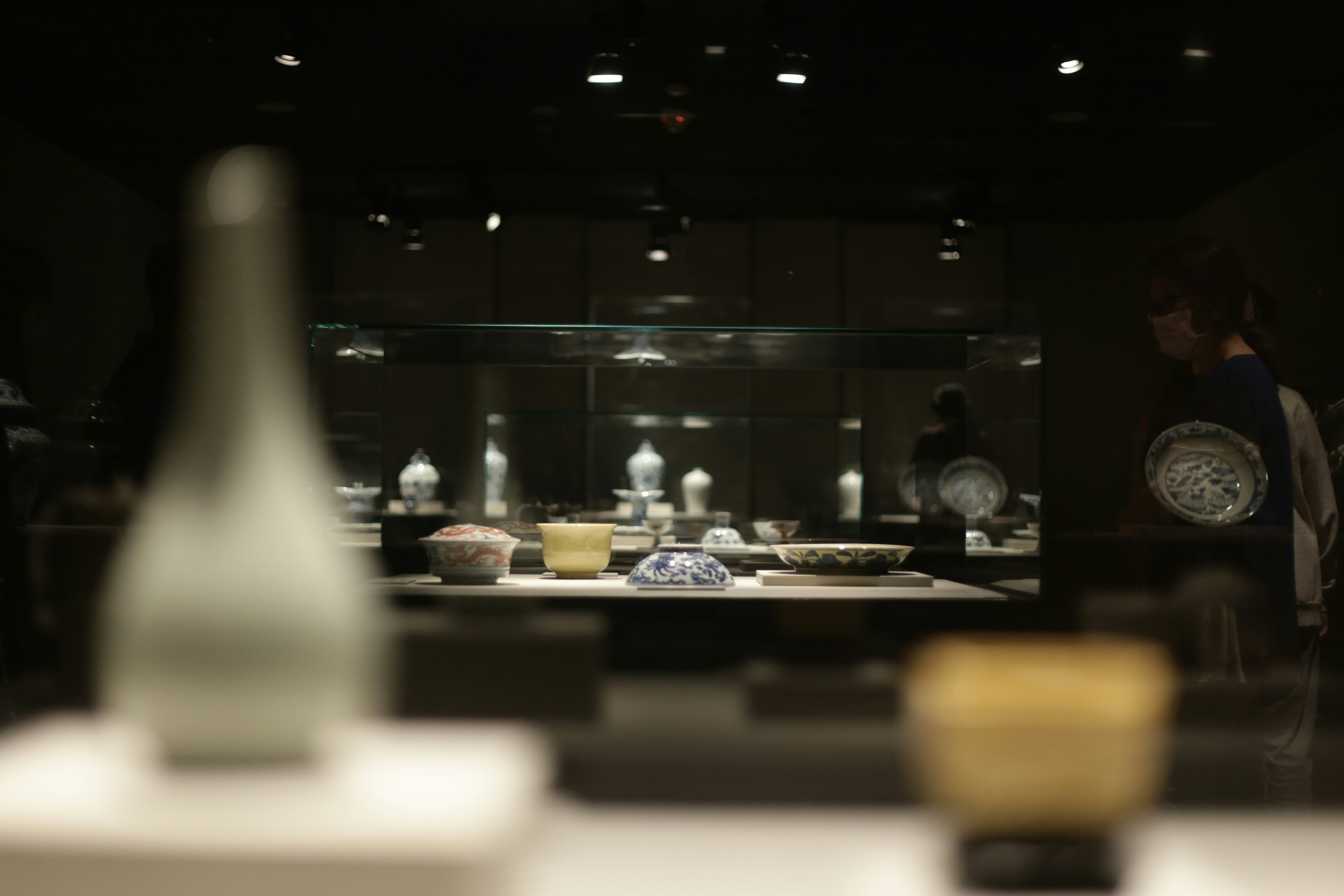 Dimly lit exhibit hall with a distant row of ceramic bowls and vases on a long display shelf; foreground features blurred pottery, conveying depth and focus on the central objects.